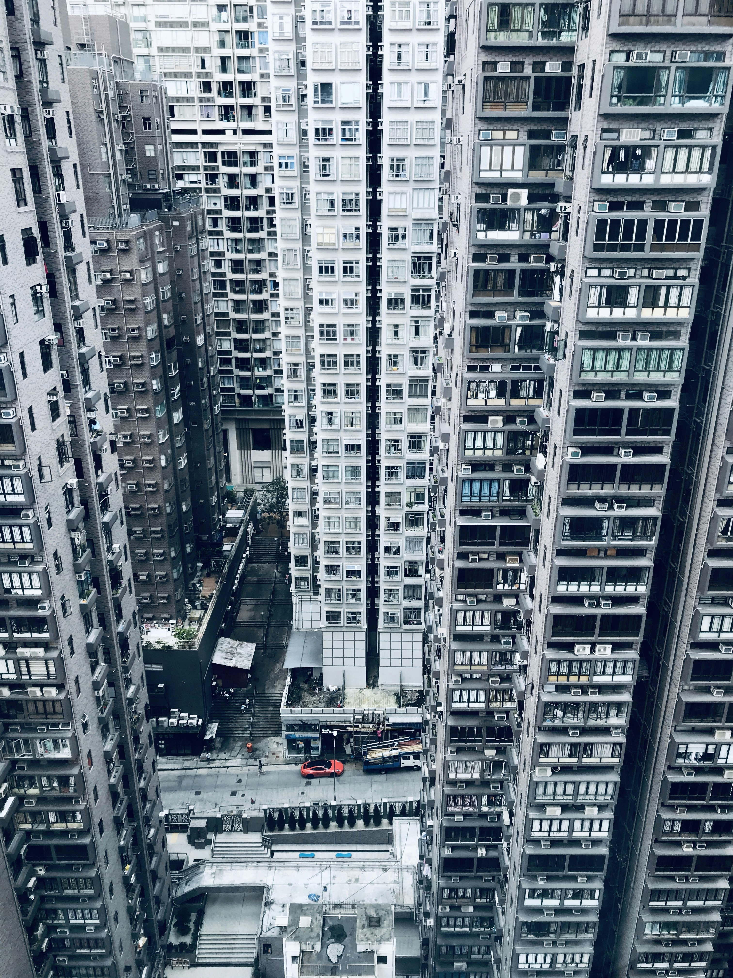 High-rise buildings create a dense urban canyon, showcasing a narrow street below with a glimpse of a red vehicle amidst the concrete structures.