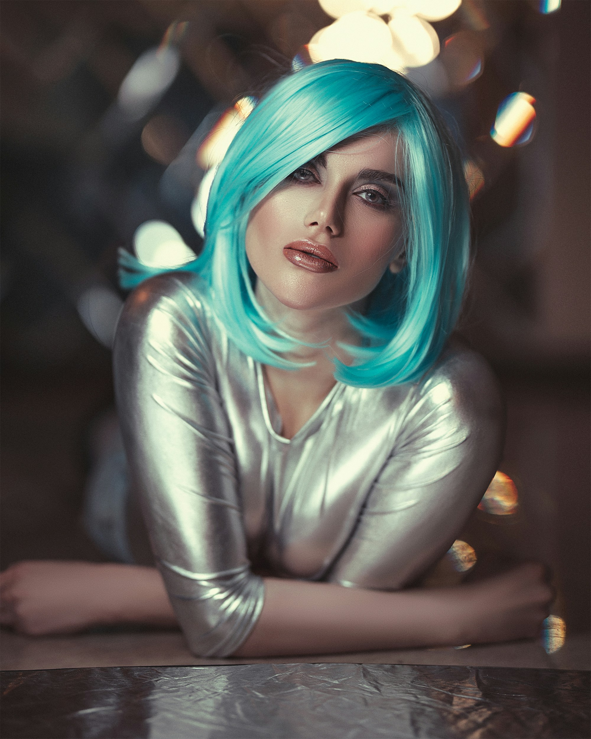 Model with turquoise hair and silver attire poses gracefully, surrounded by soft bokeh lights, creating a dreamlike atmosphere.