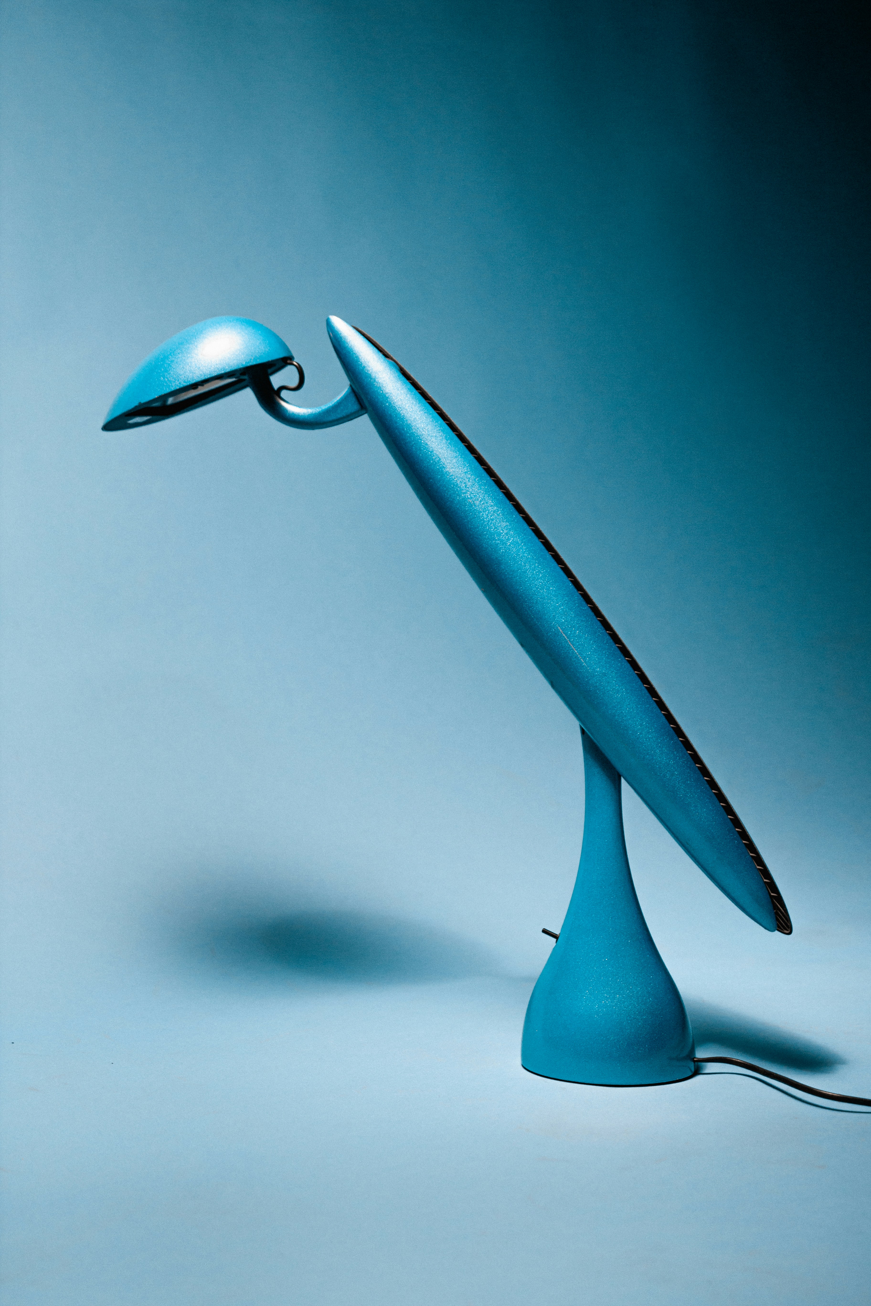 Blue desk lamp with a curved neck rests on a smooth surface against a cool blue backdrop, its form simplified into a sculptural silhouette.