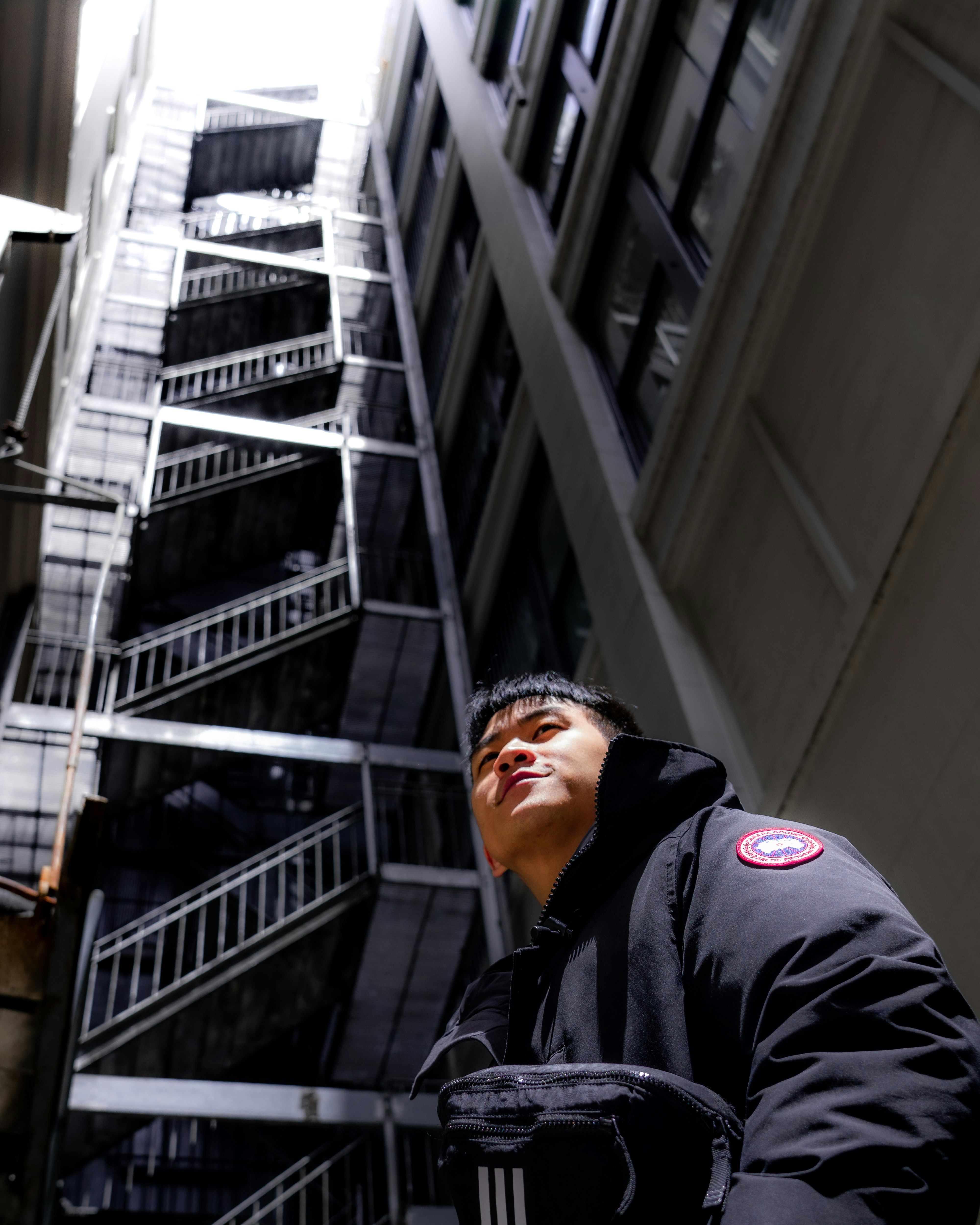Man gazing upward in a narrow alley flanked by towering buildings and a fire escape. The scene captures the essence of urban life and architectural contrasts.