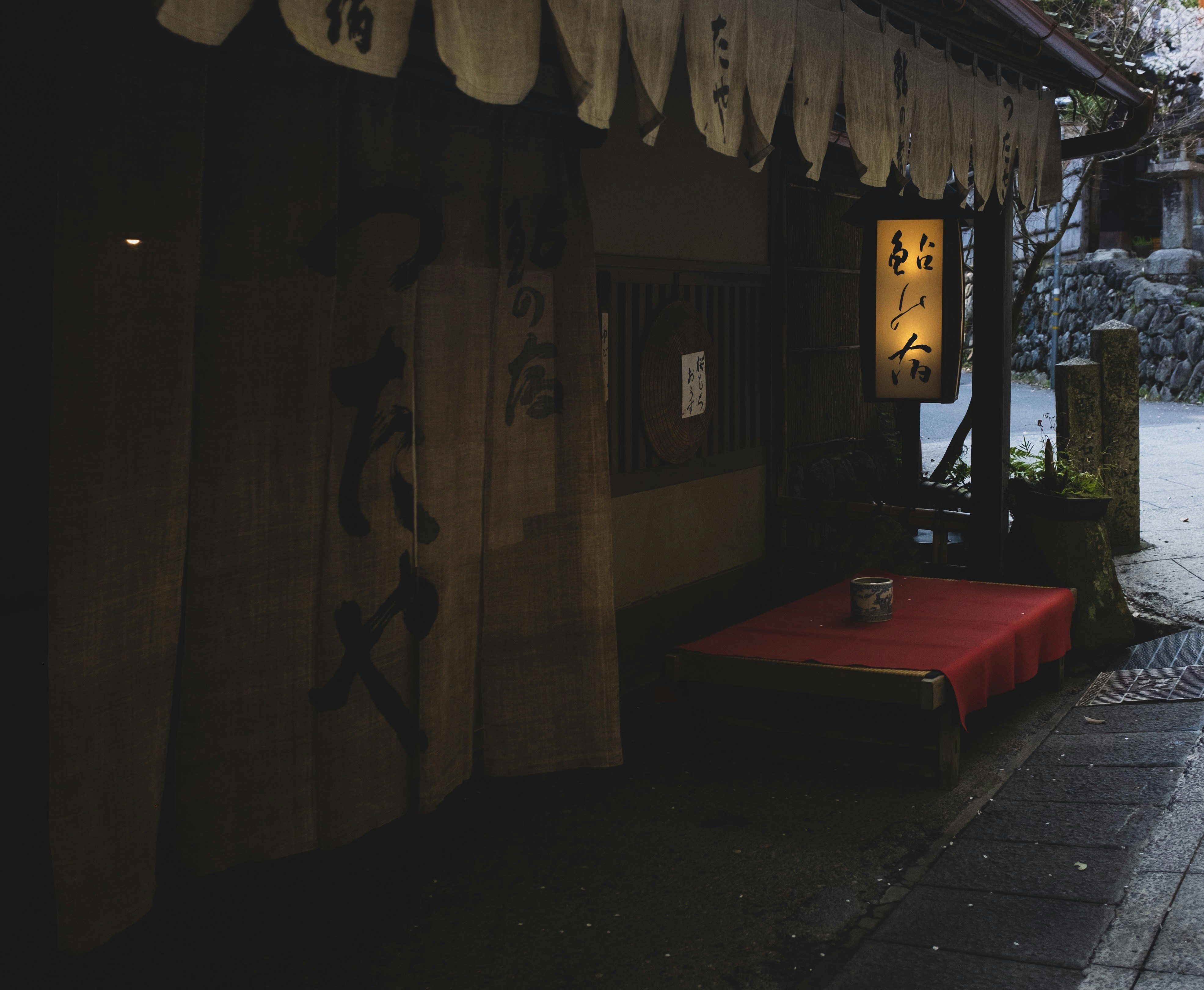 A cozy Japanese eatery adorned with hanging fabric signage, featuring a red table set outside, inviting passersby to experience local cuisine.