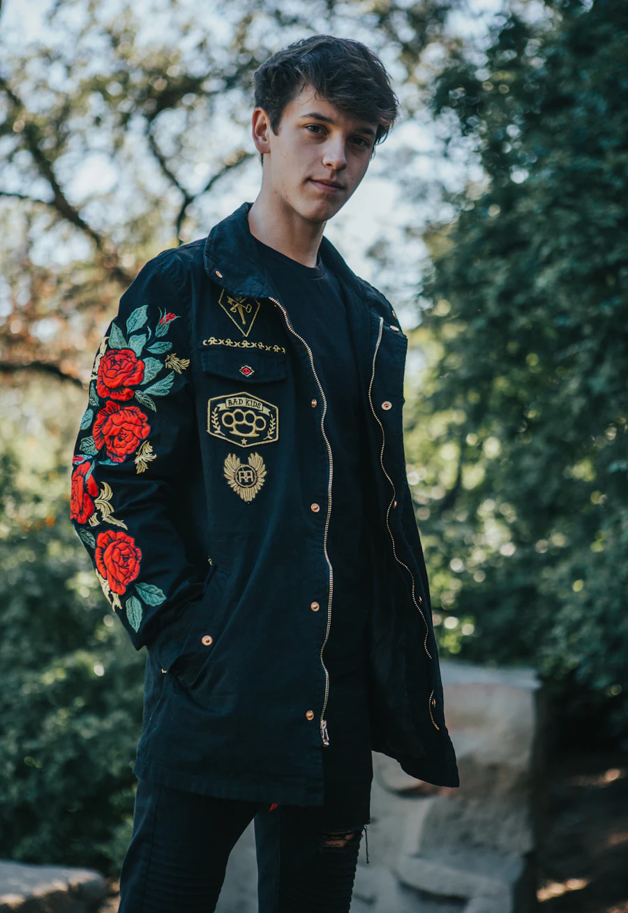 Young man standing outdoors in a black jacket with red rose embroidery on the sleeves, looking toward the camera with trees blurred behind him.