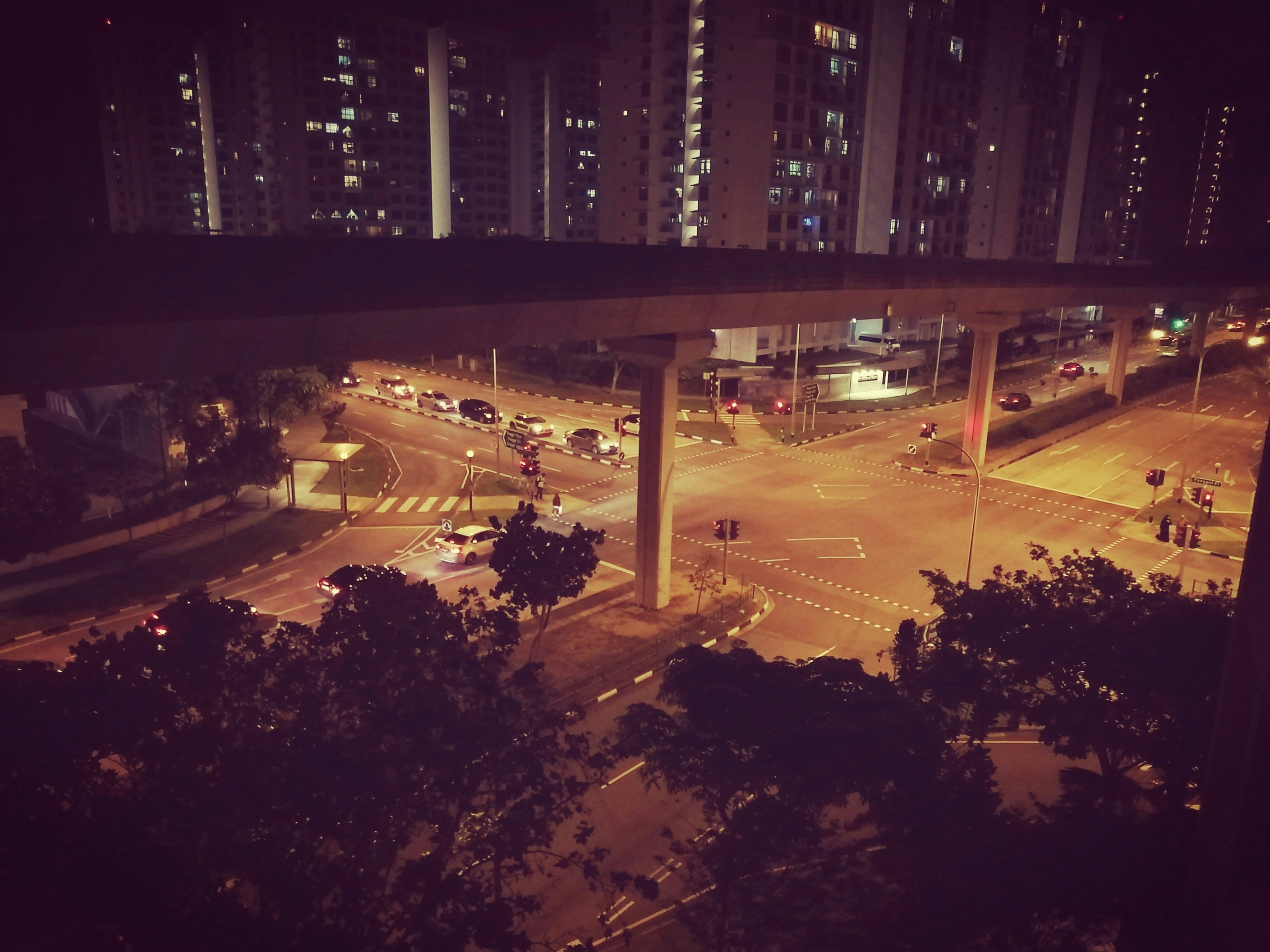Elevated highway crossing above a bustling city intersection at night, framed by tall buildings.