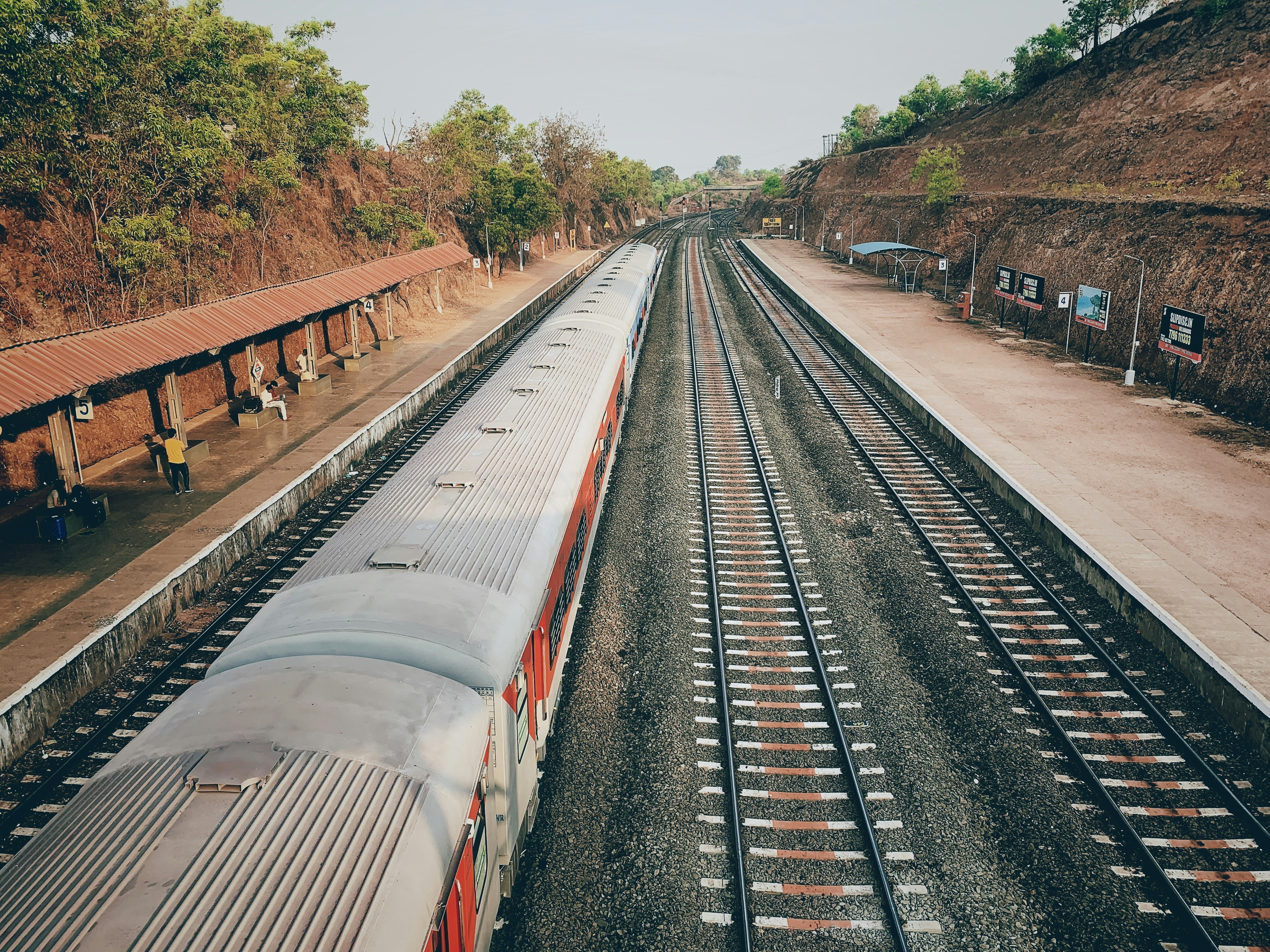 A train glides along empty tracks at a tranquil station, framed by lush greenery and a distant hillside. The scene captures the essence of travel and solitude.