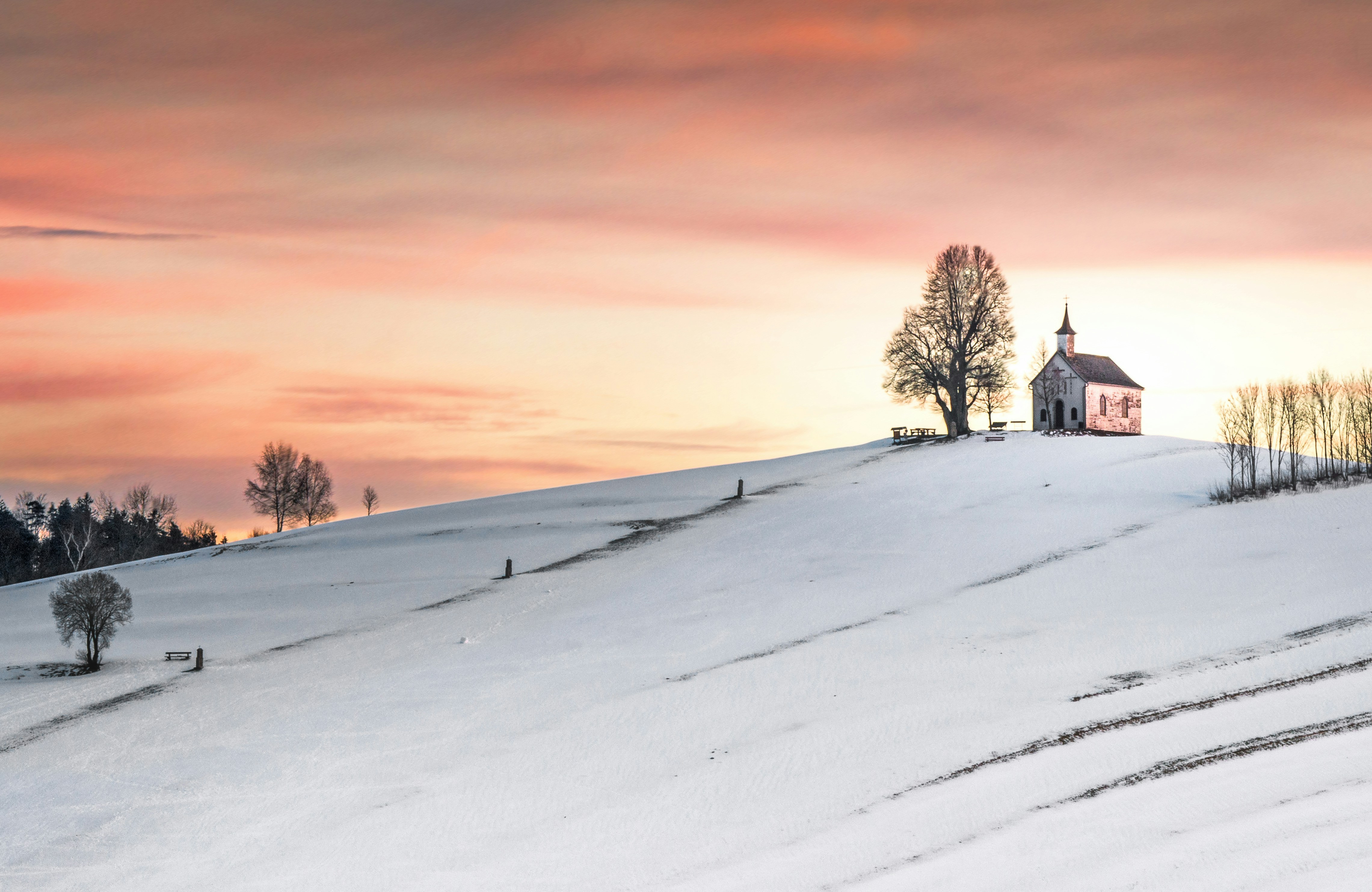 A quaint chapel stands alone on a snowy hill under a pastel sky at dusk. The serene landscape captures the tranquility of winter.