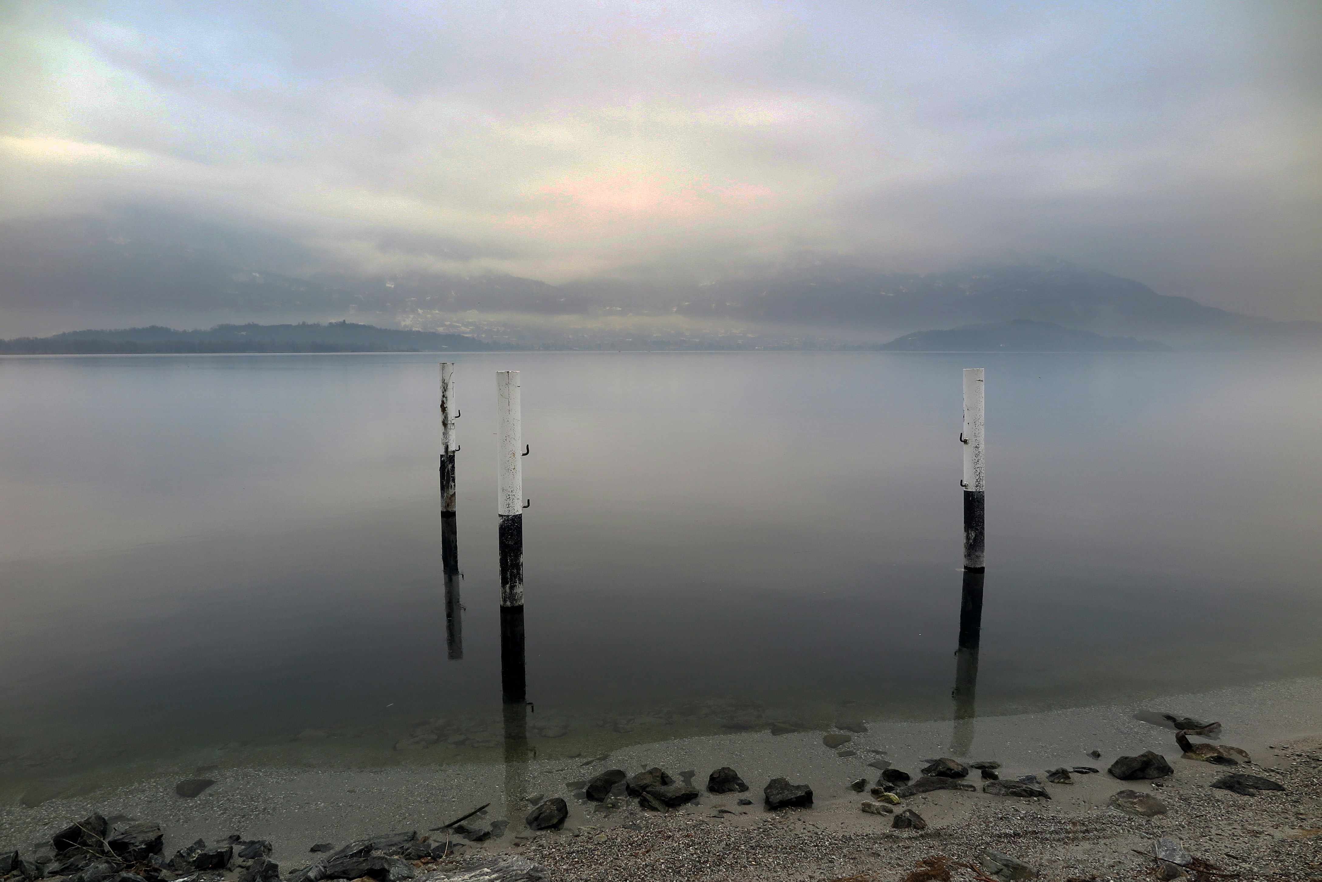 Three wooden poles stand in calm water beneath a cloudy sky, mountains faintly visible in the distance.