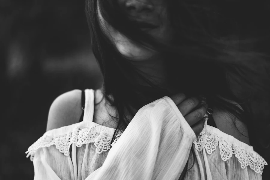 Black-and-white portrait of a woman with windblown hair, wearing a lace-trimmed off-shoulder blouse and holding fabric near her chest. Her face is partly obscured, creating a moody, intimate feel.