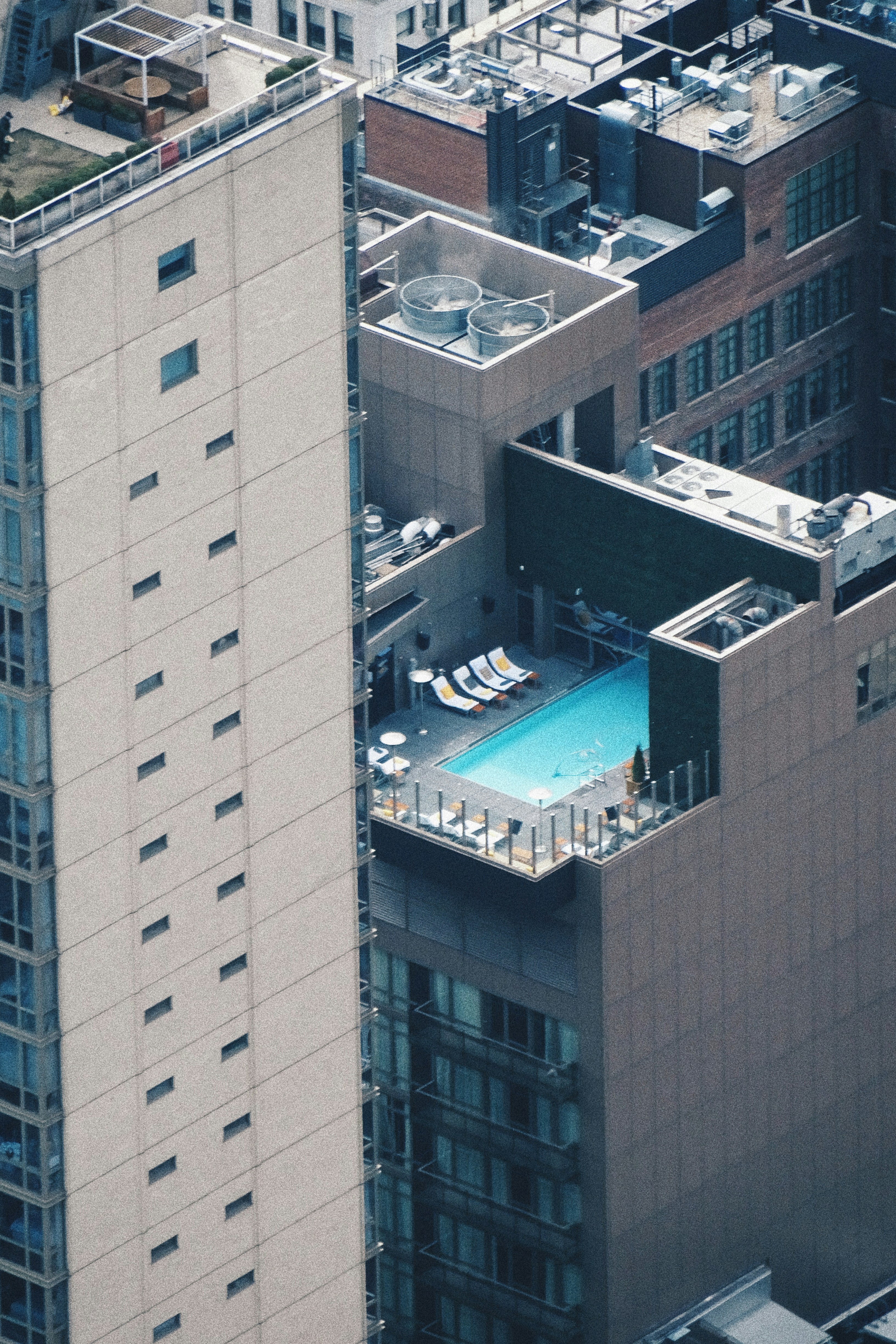 Rooftop swimming pool flanked by high-rise buildings, viewed from above. A compact urban leisure space framed by concrete towers.