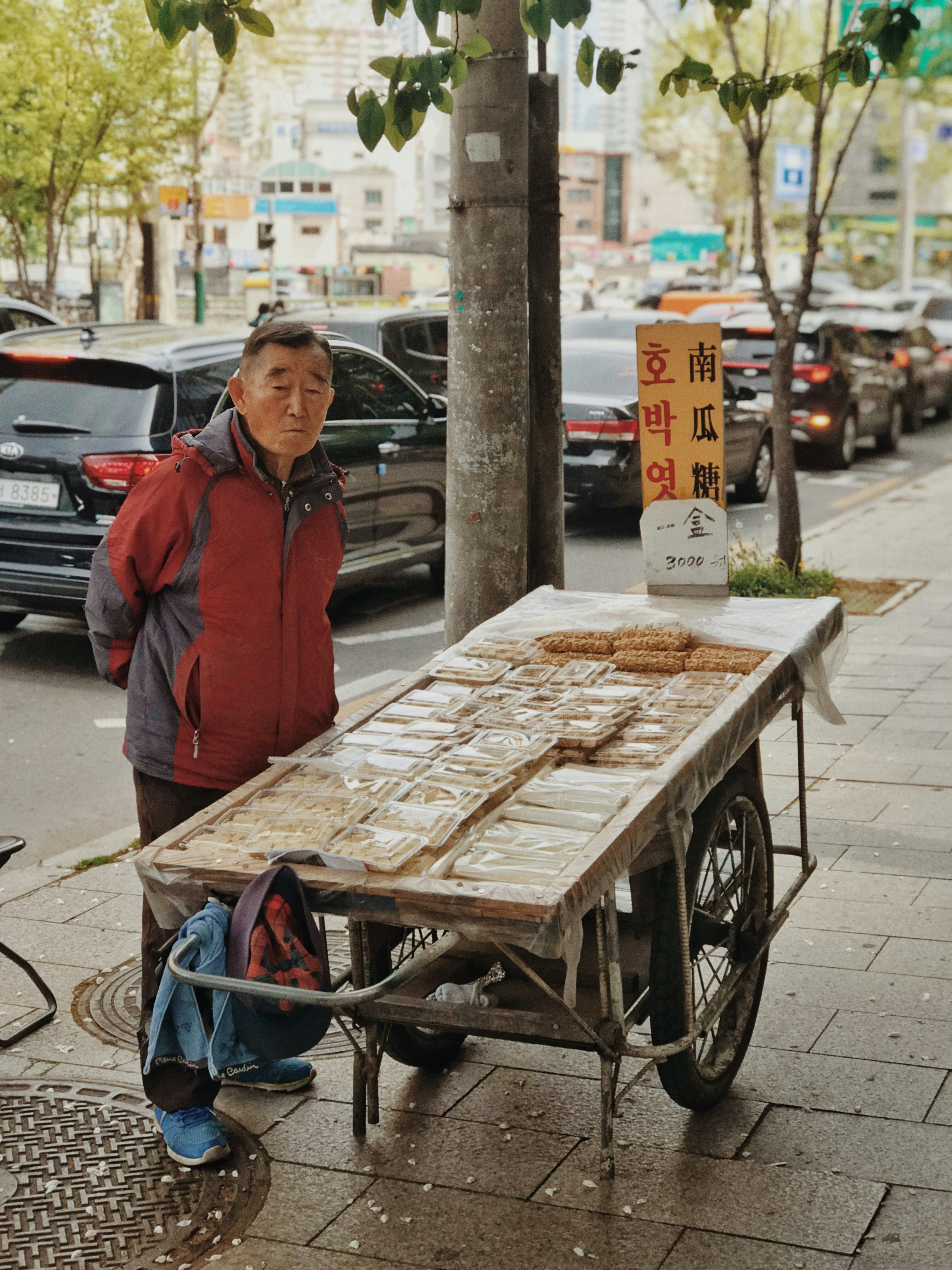 Red-Jacket Vendor Beside a Wheeled Cart of Wrapped Goods on a Busy City Street