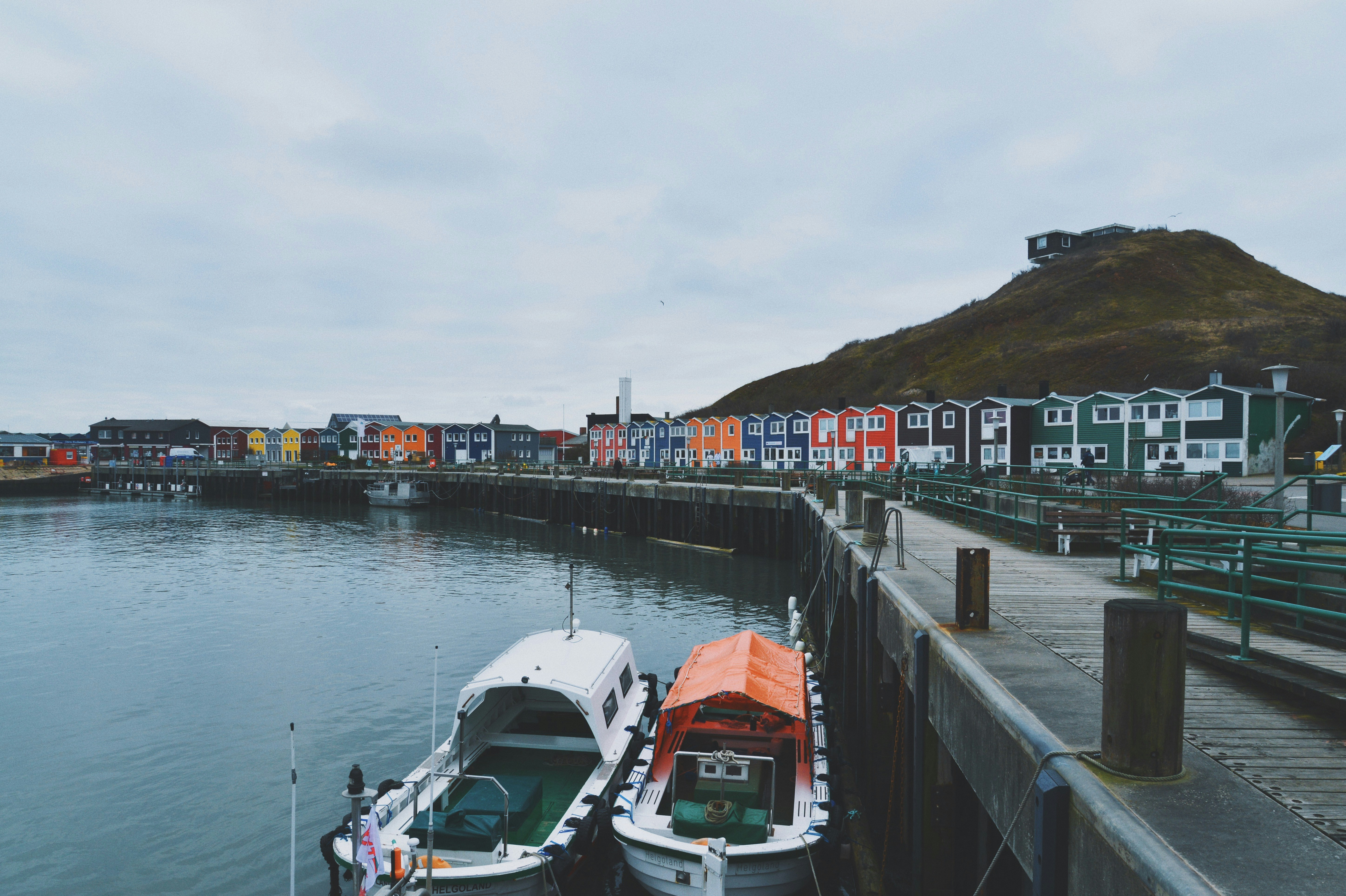 Colorful houses line the harbor, reflecting in the calm waters, while boats rest along the dock. A hillside lookout completes the picturesque scene.