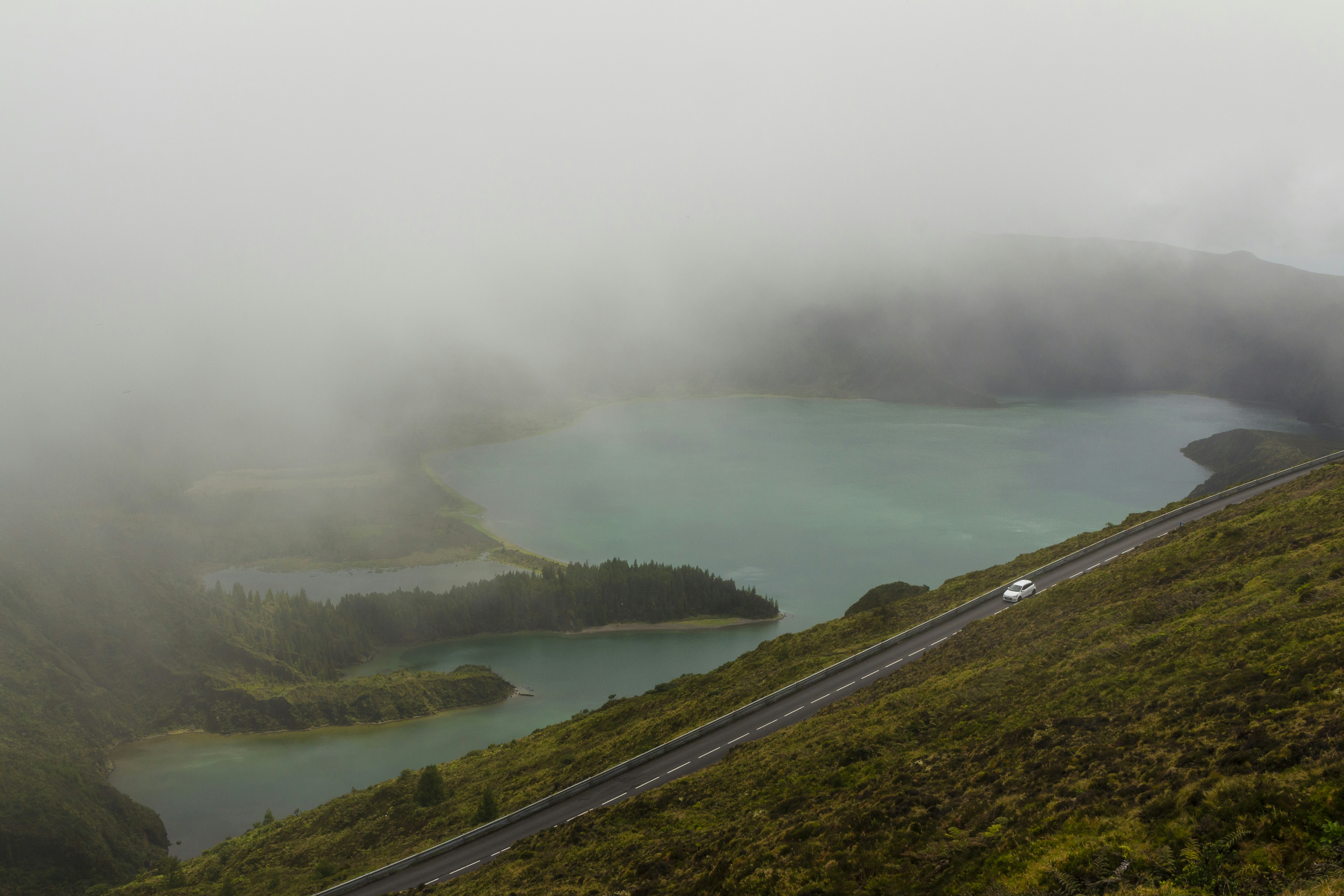 Fog drifts over a tranquil lake bordered by lush hills and a winding road.