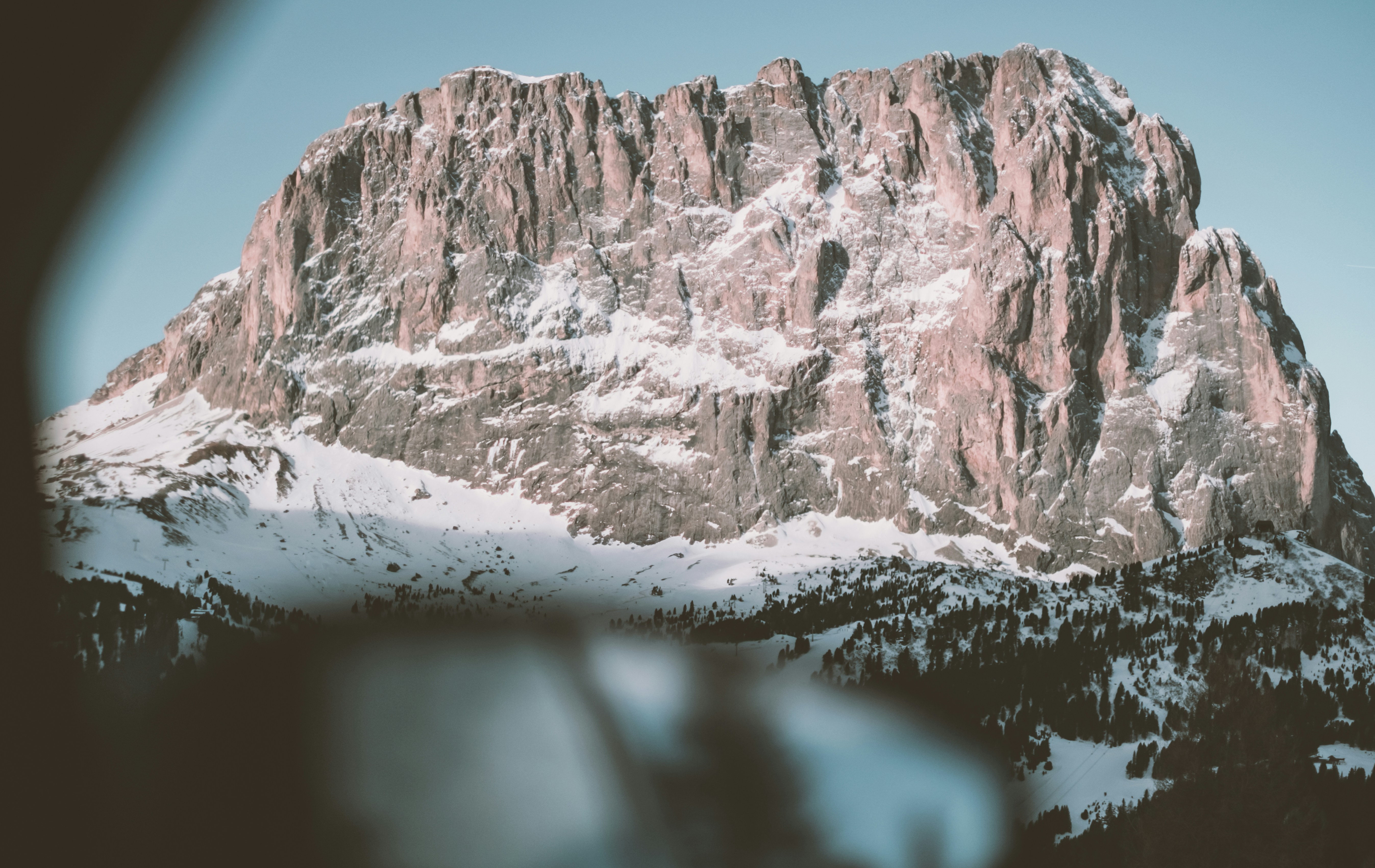 Snow-capped Saslonch mountain under a soft blue sky, framed by blurred foreground elements.