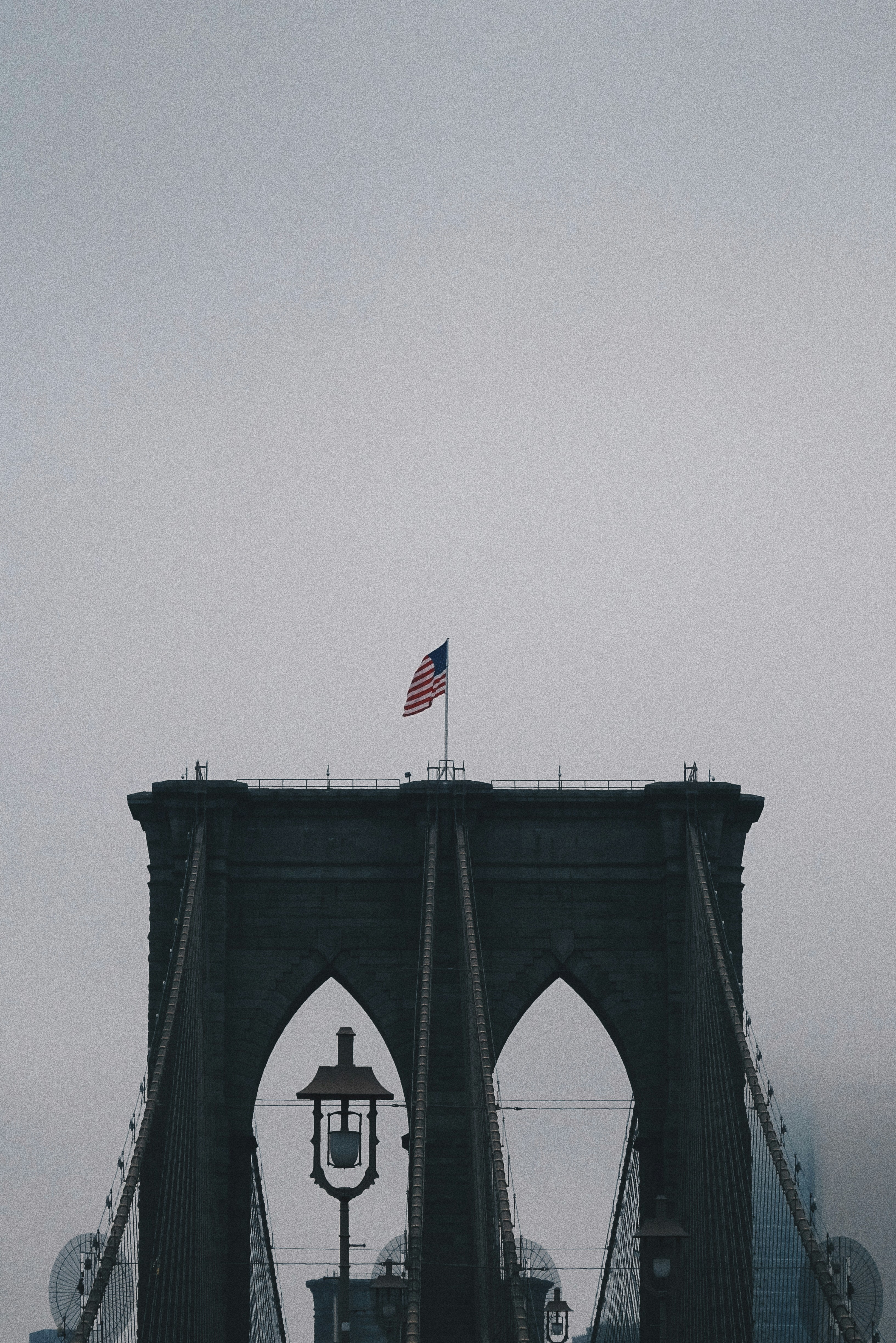 The American flag waves atop the Brooklyn Bridge, framed by its iconic arches and a moody skyline. The atmosphere evokes a sense of history and resilience.