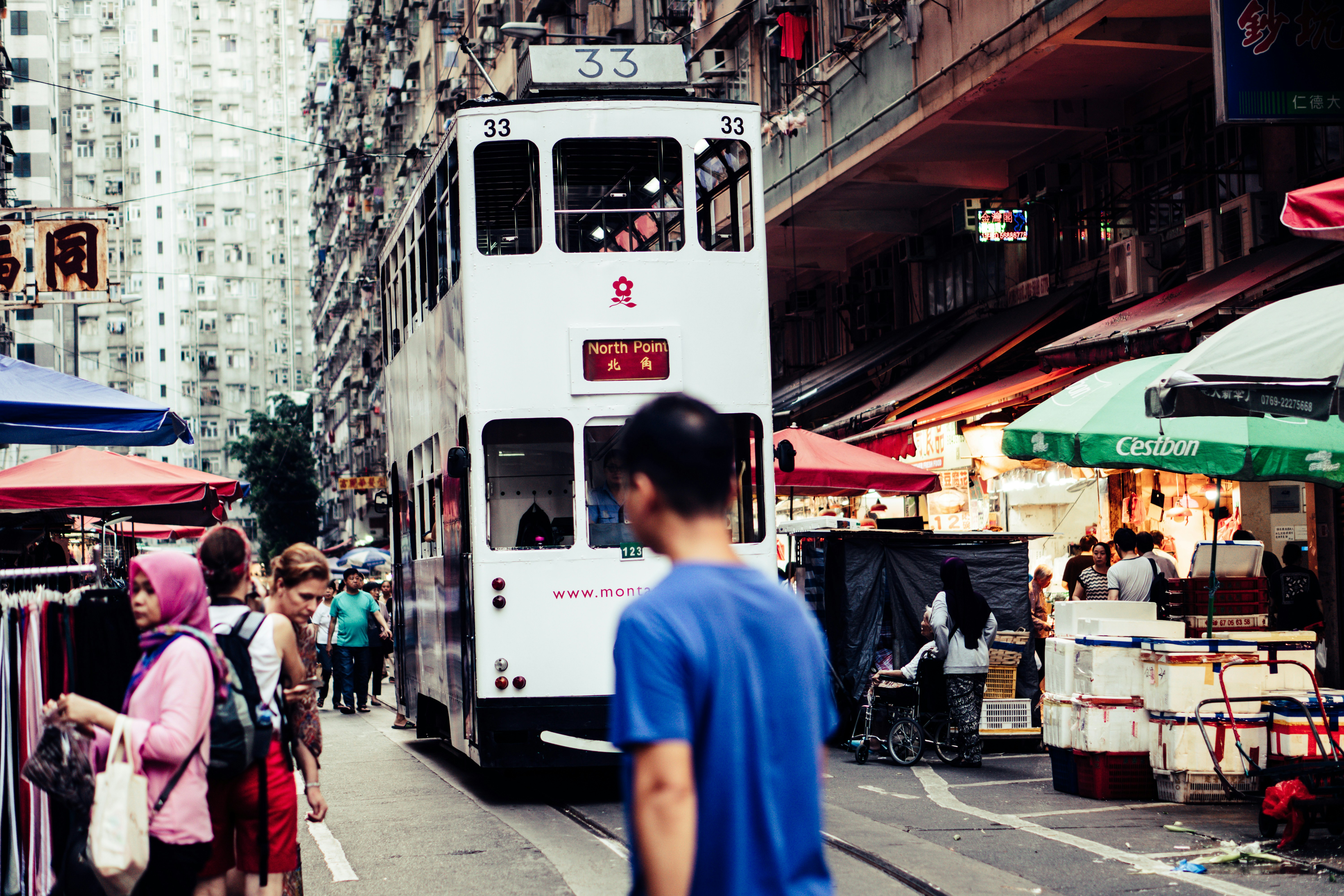 A vintage tram navigates a bustling street market, surrounded by vibrant stalls and pedestrians. The scene captures the essence of urban life in a lively city.