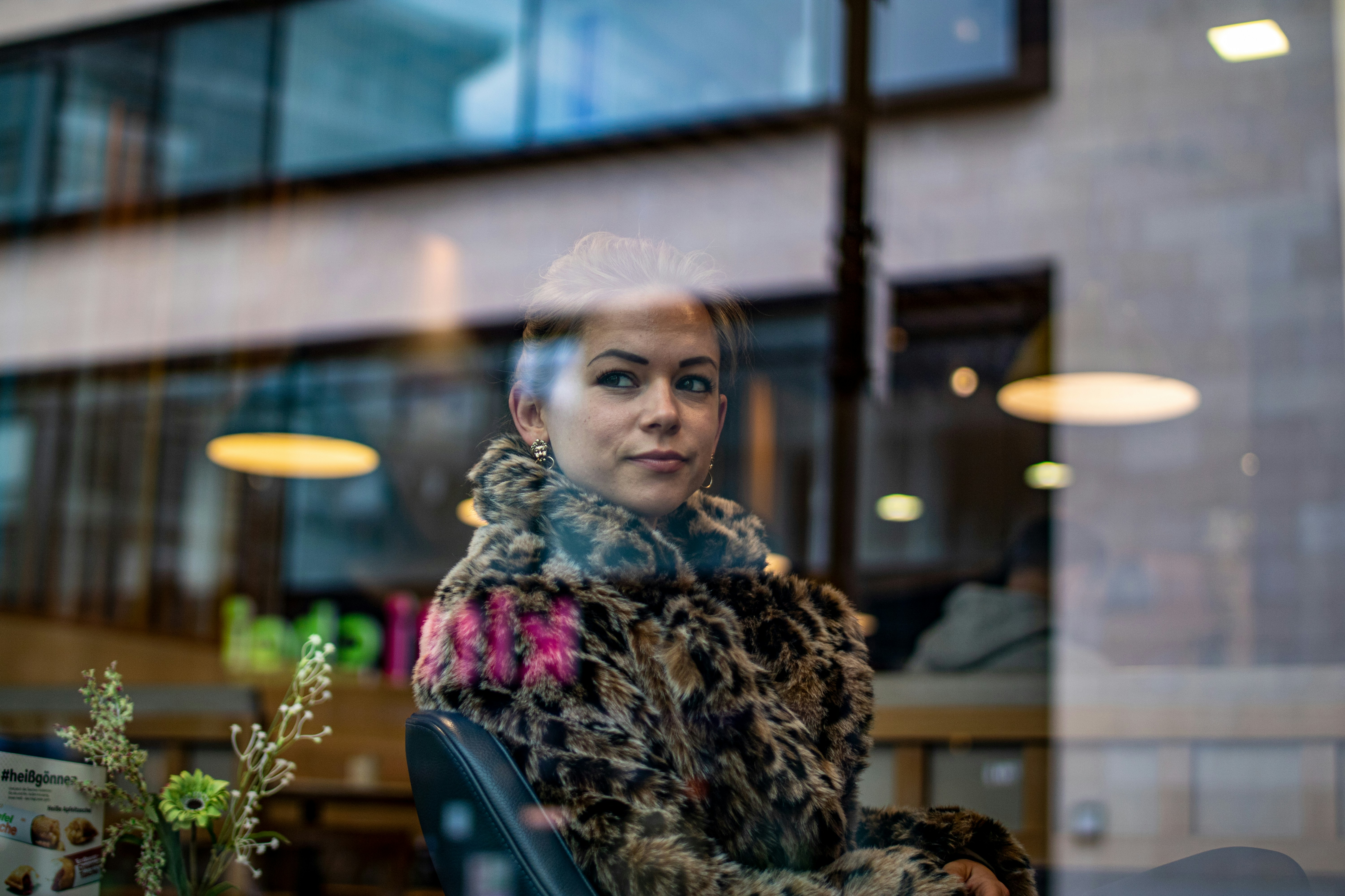 A young woman in a stylish fur coat gazes thoughtfully through a café window, reflecting the ambient light and interior decor.