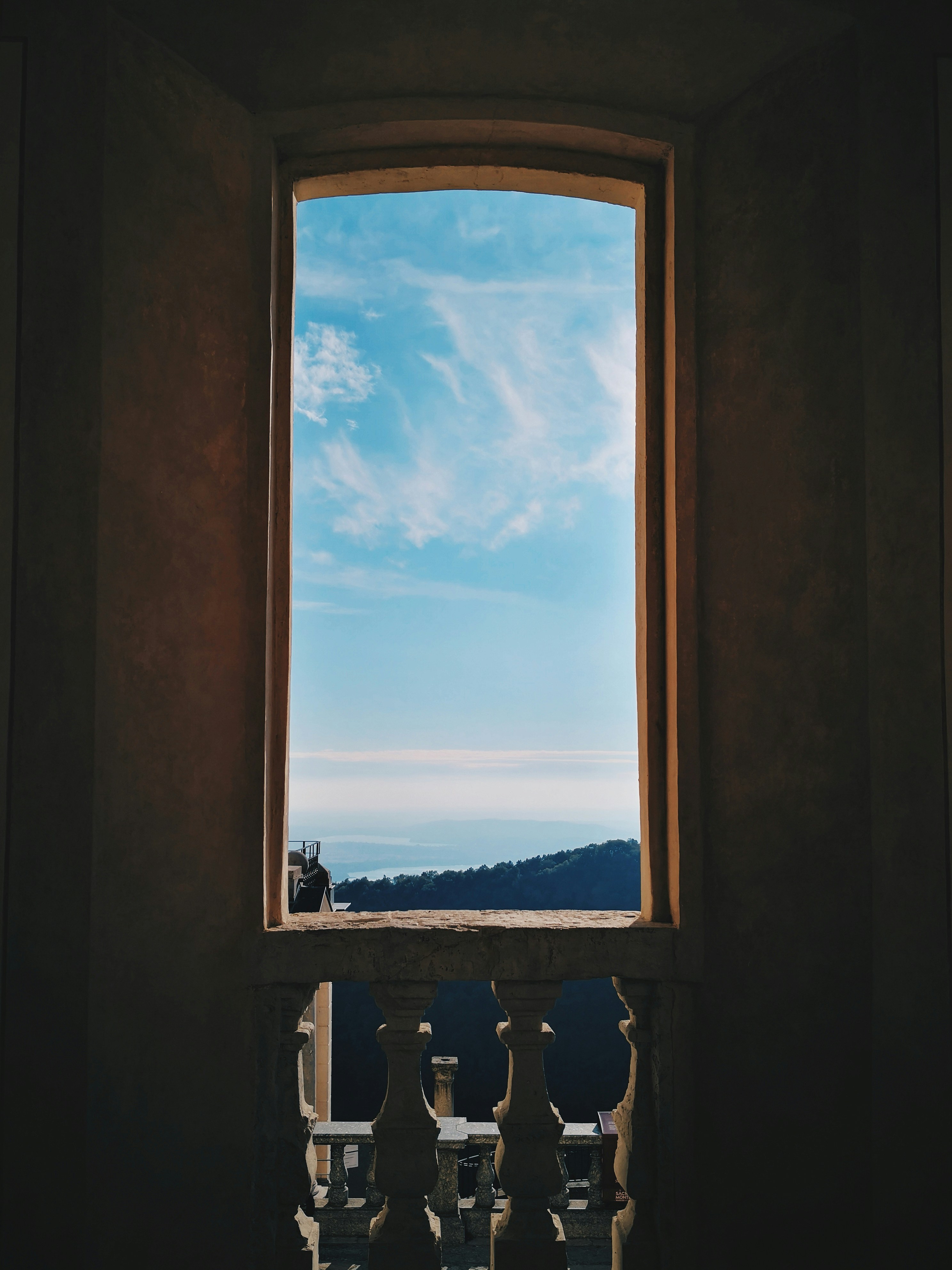 Dark interior frames a tall window opening onto a blue sky and distant sea, with a stone balustrade in the foreground.
