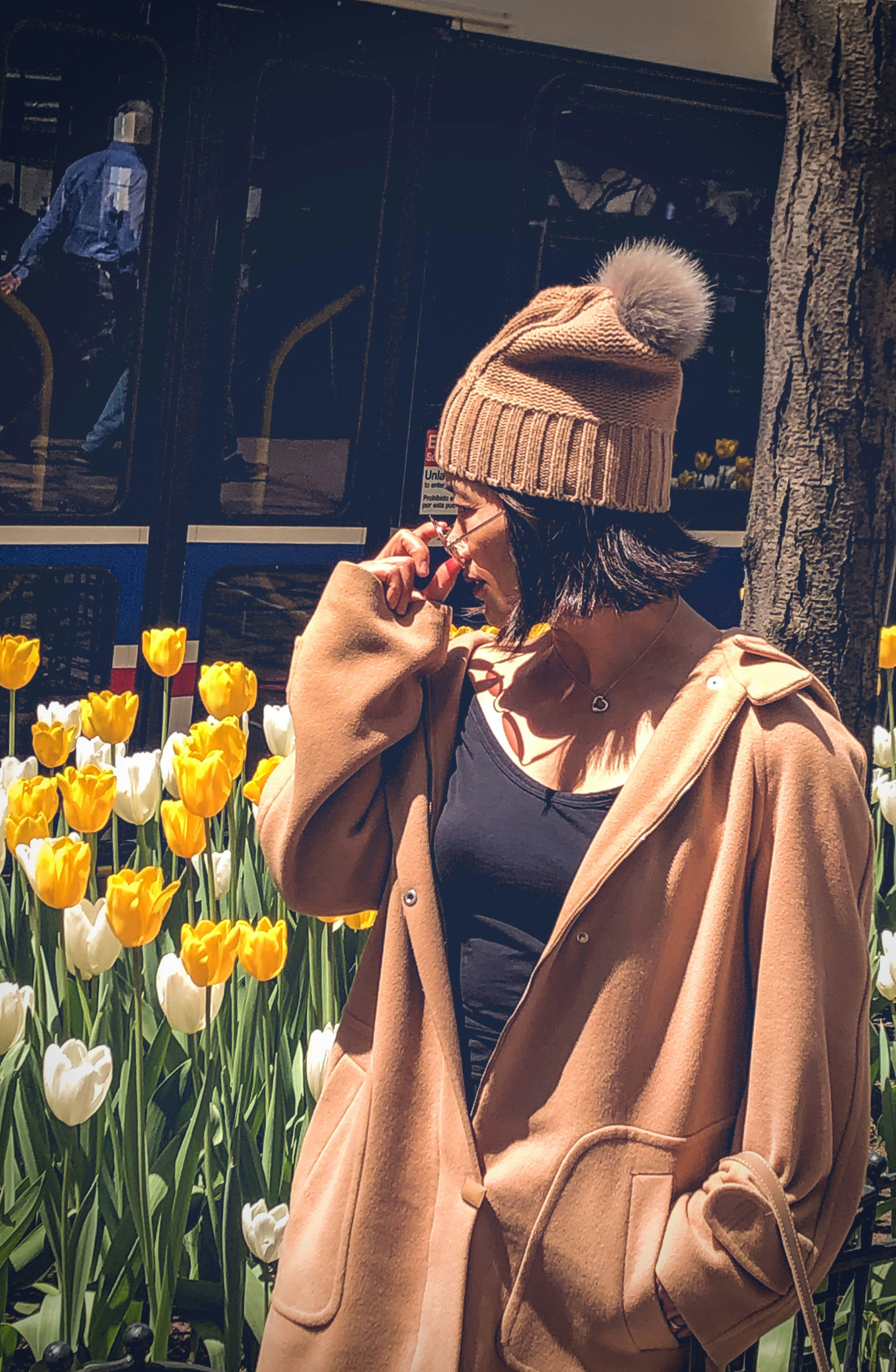 Woman in a brown coat and hat stands by blooming yellow and white tulips.