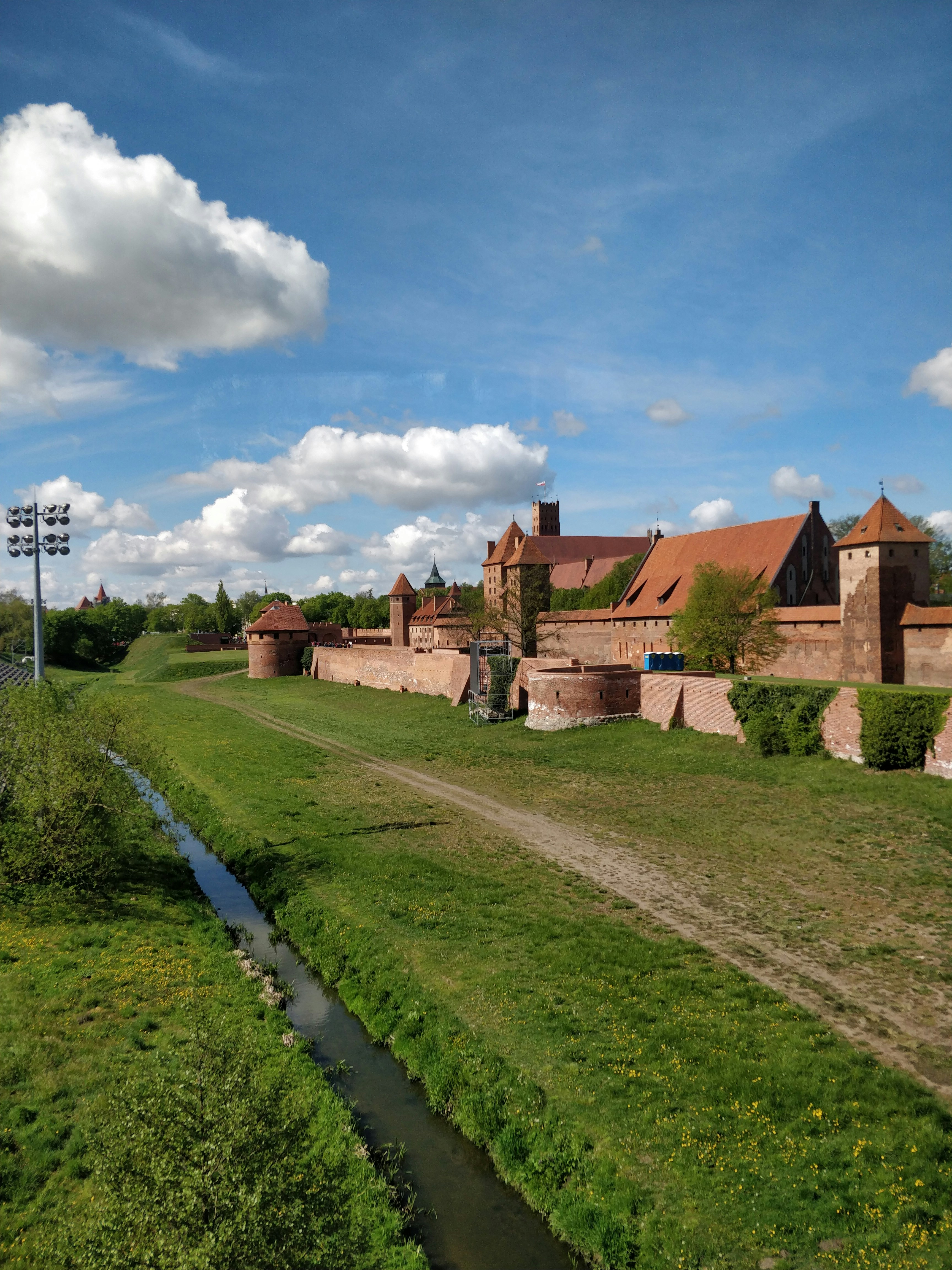 Historic brick fortress with a lush green landscape and a winding stream in the foreground, under a blue sky dotted with clouds.