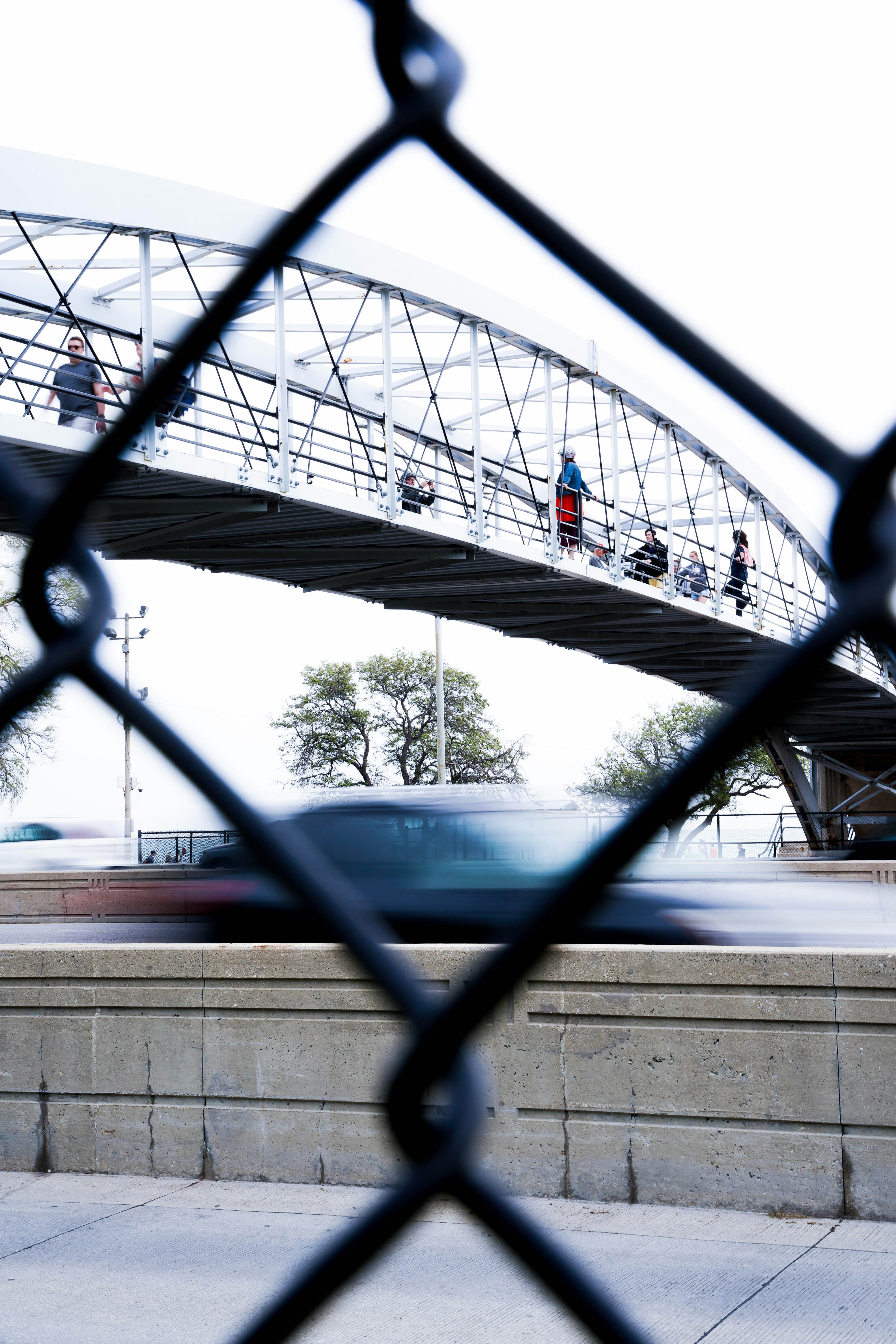 Traffic on a Curved Footbridge, Framed by Chain-Link Silhouette