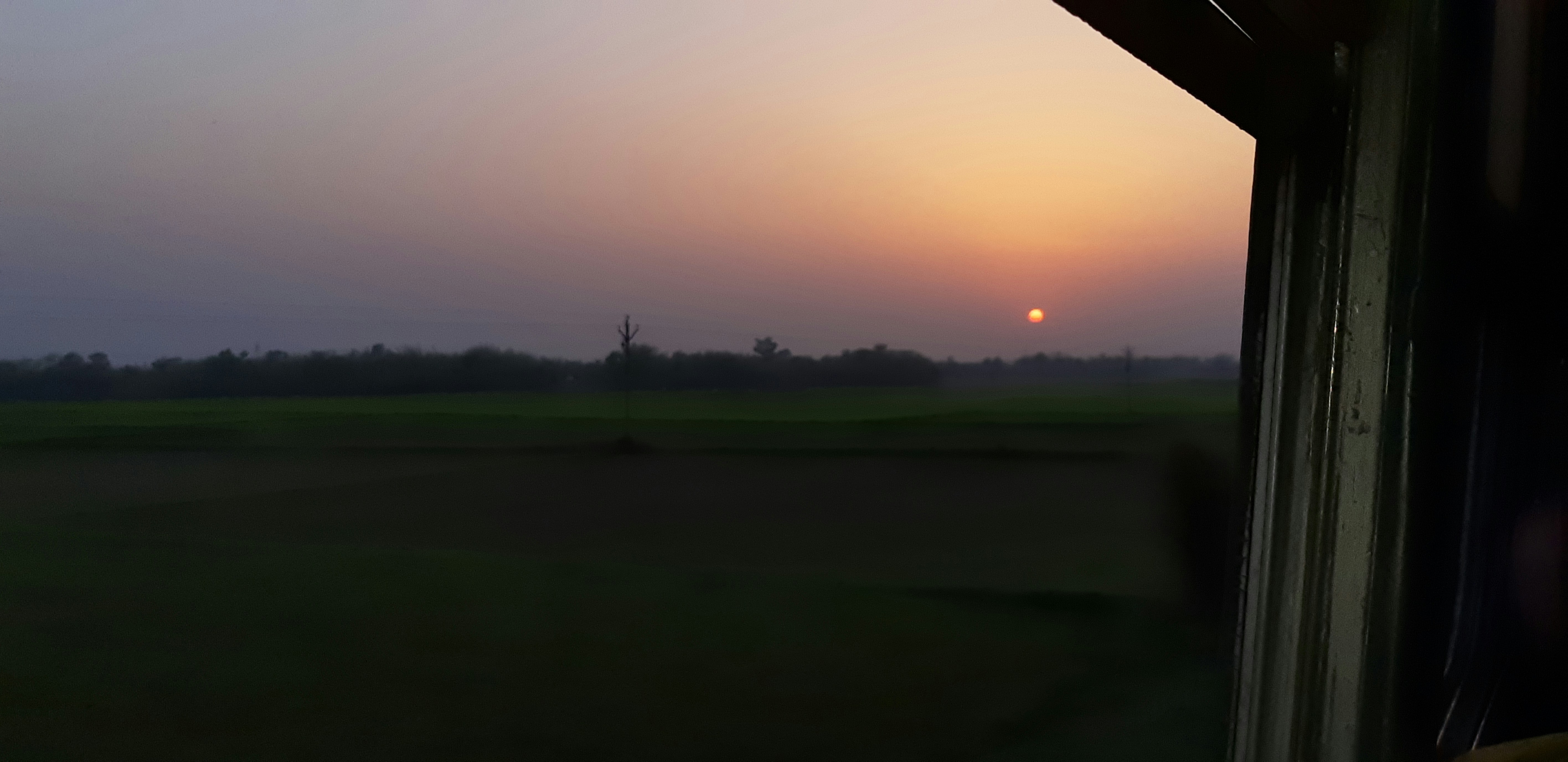 The sun sets behind lush fields, casting a warm glow across the horizon. The scene captures the tranquility of rural life at twilight.
