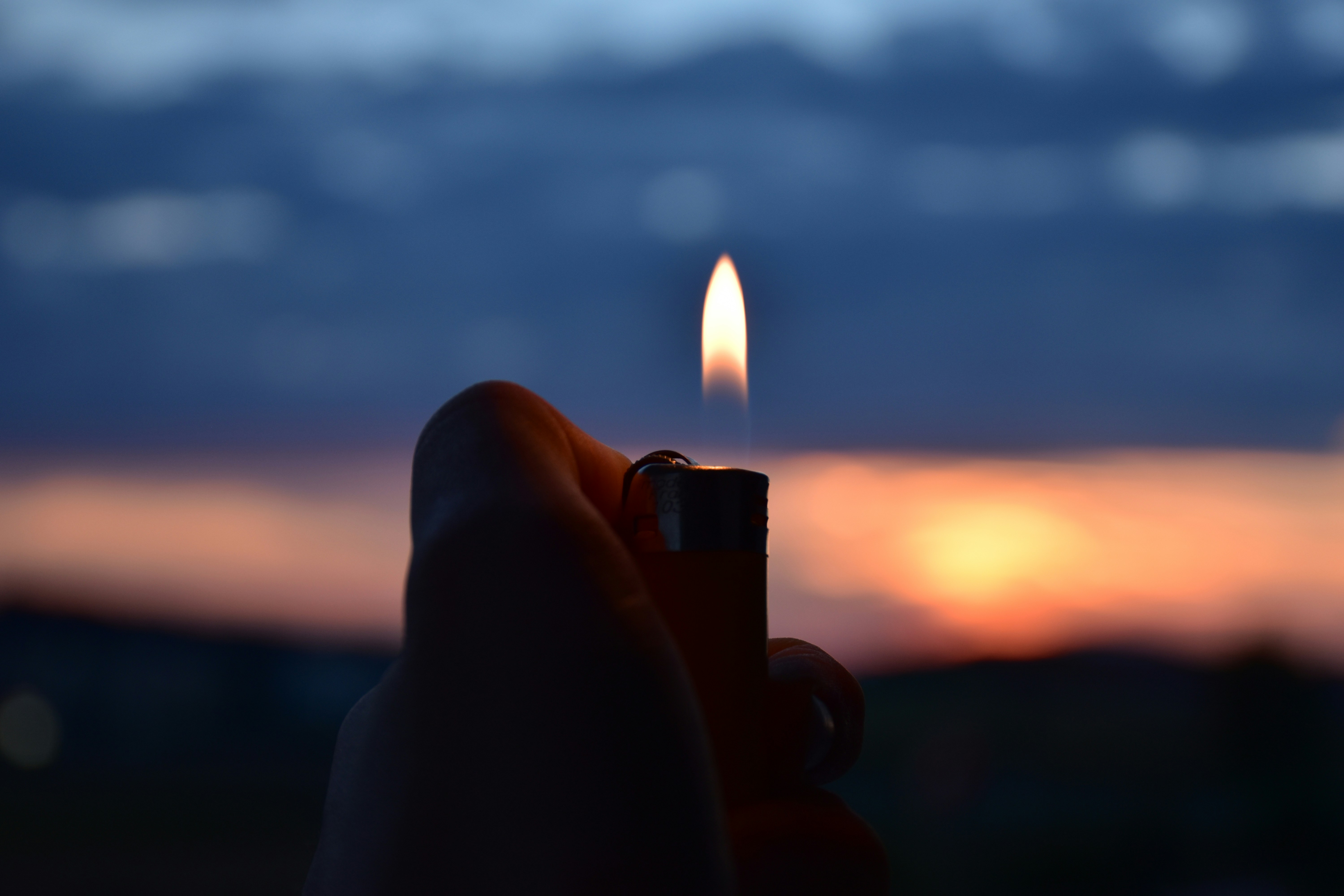 A hand holding a lighter with a vibrant flame, silhouetted against a colorful sunset sky.