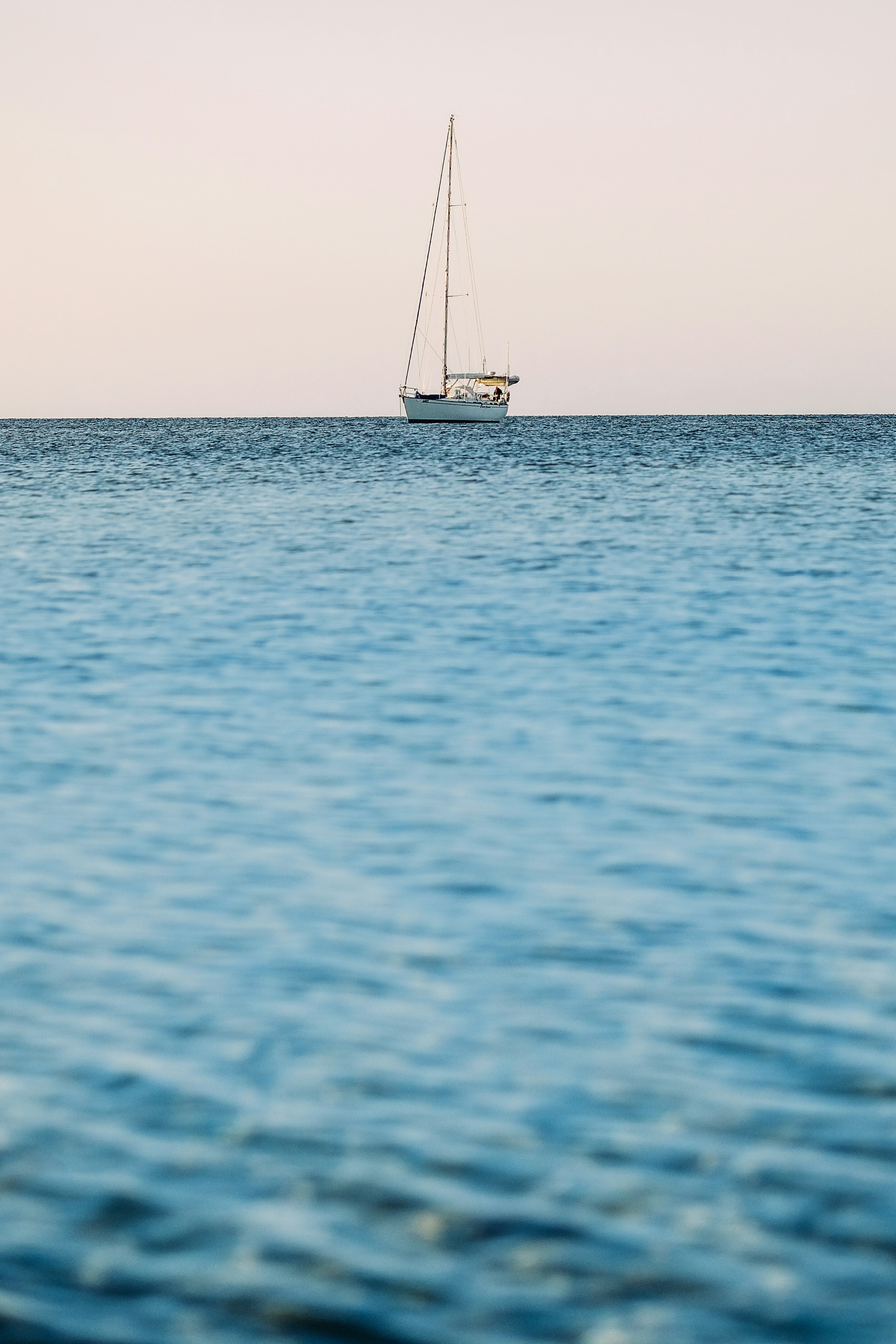A solitary sailboat glides across the calm sea, with a soft pastel sky reflecting on the water's surface.