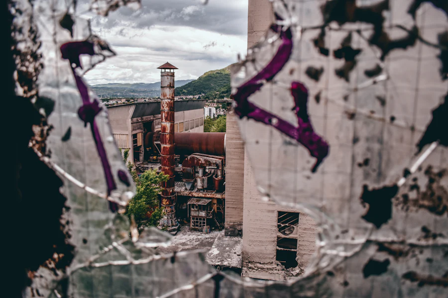 Industrial ruins viewed through broken glass and barbed wire, with distant hills and a weathered smokestack creating depth and contrast.