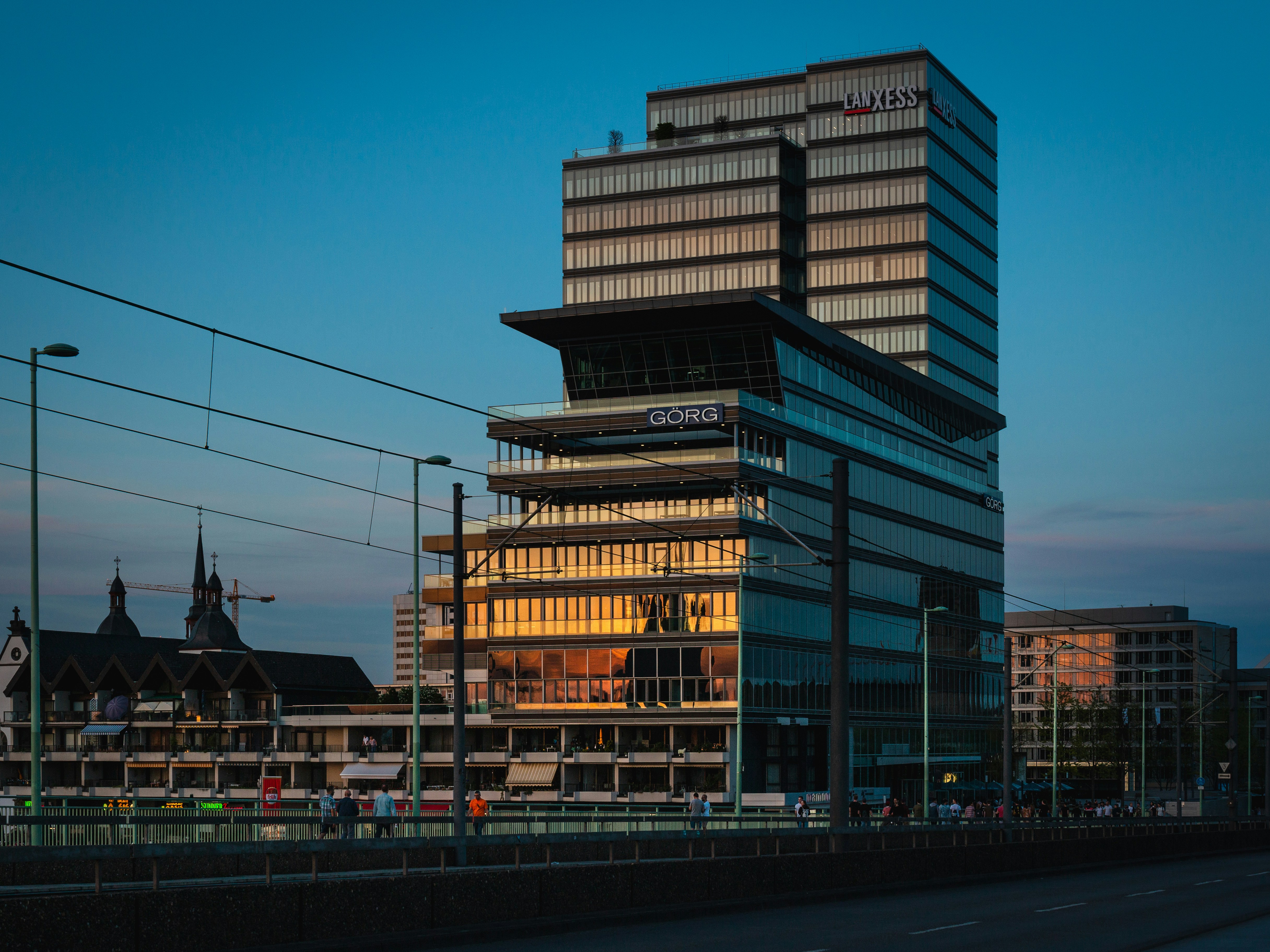 Modern glass-paneled building reflecting warm sunset hues with people walking on a nearby bridge.