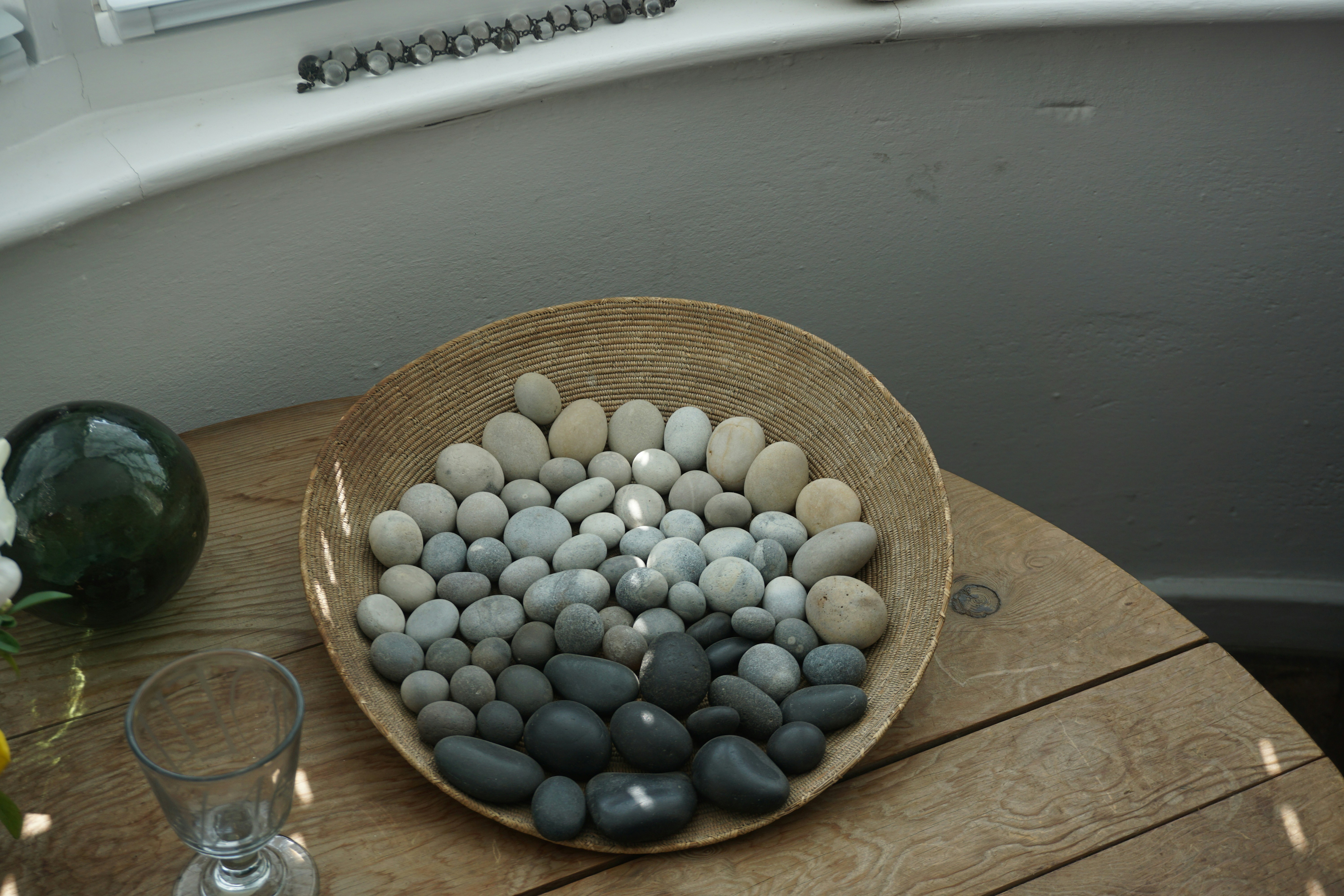 A woven bowl filled with a variety of smooth, rounded stones in shades of gray and white, arranged artfully on a wooden table.