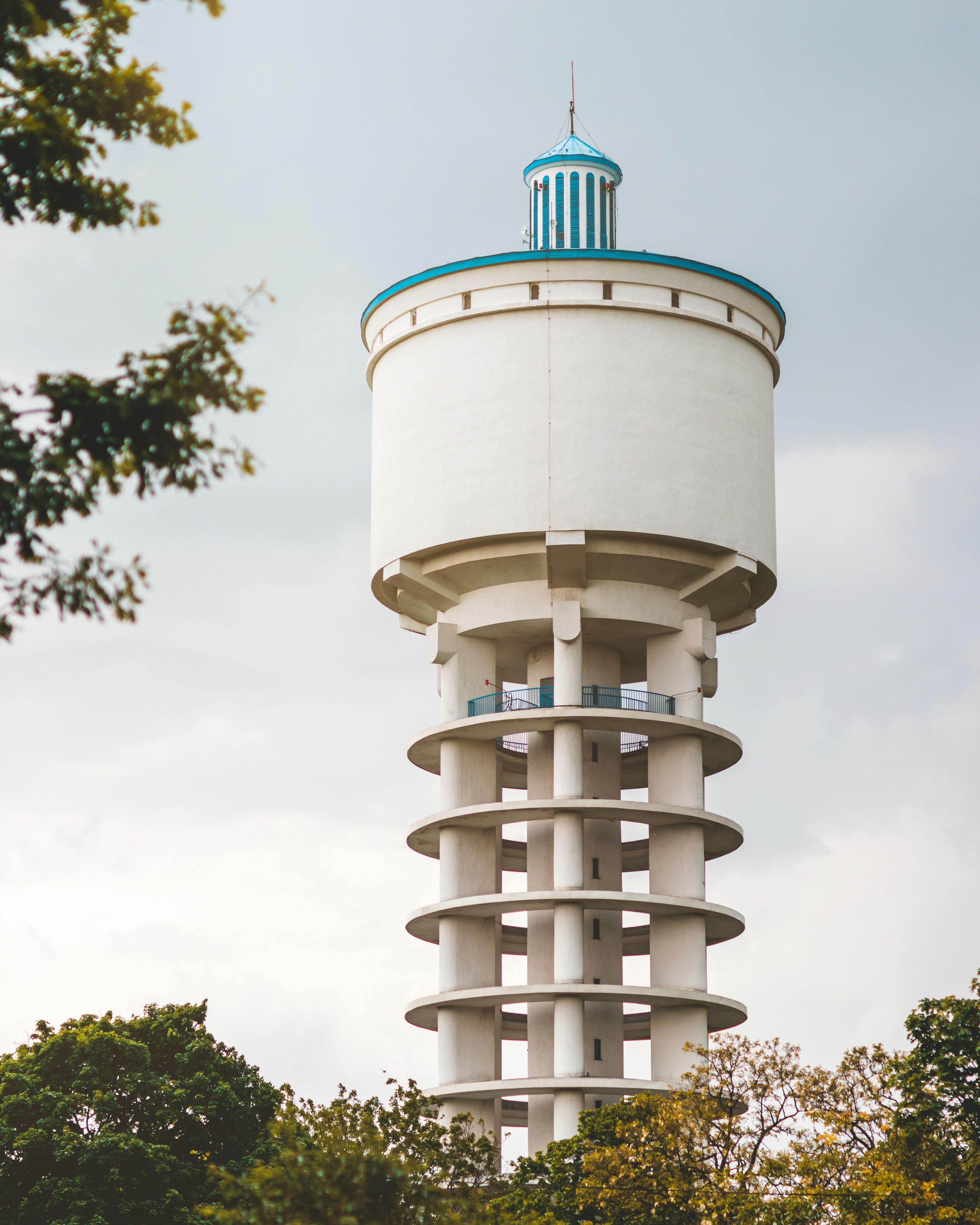 A sleek, modern water tower stands tall against a cloudy sky, surrounded by lush greenery. Its unique design features a cylindrical body and a distinctively shaped roof.