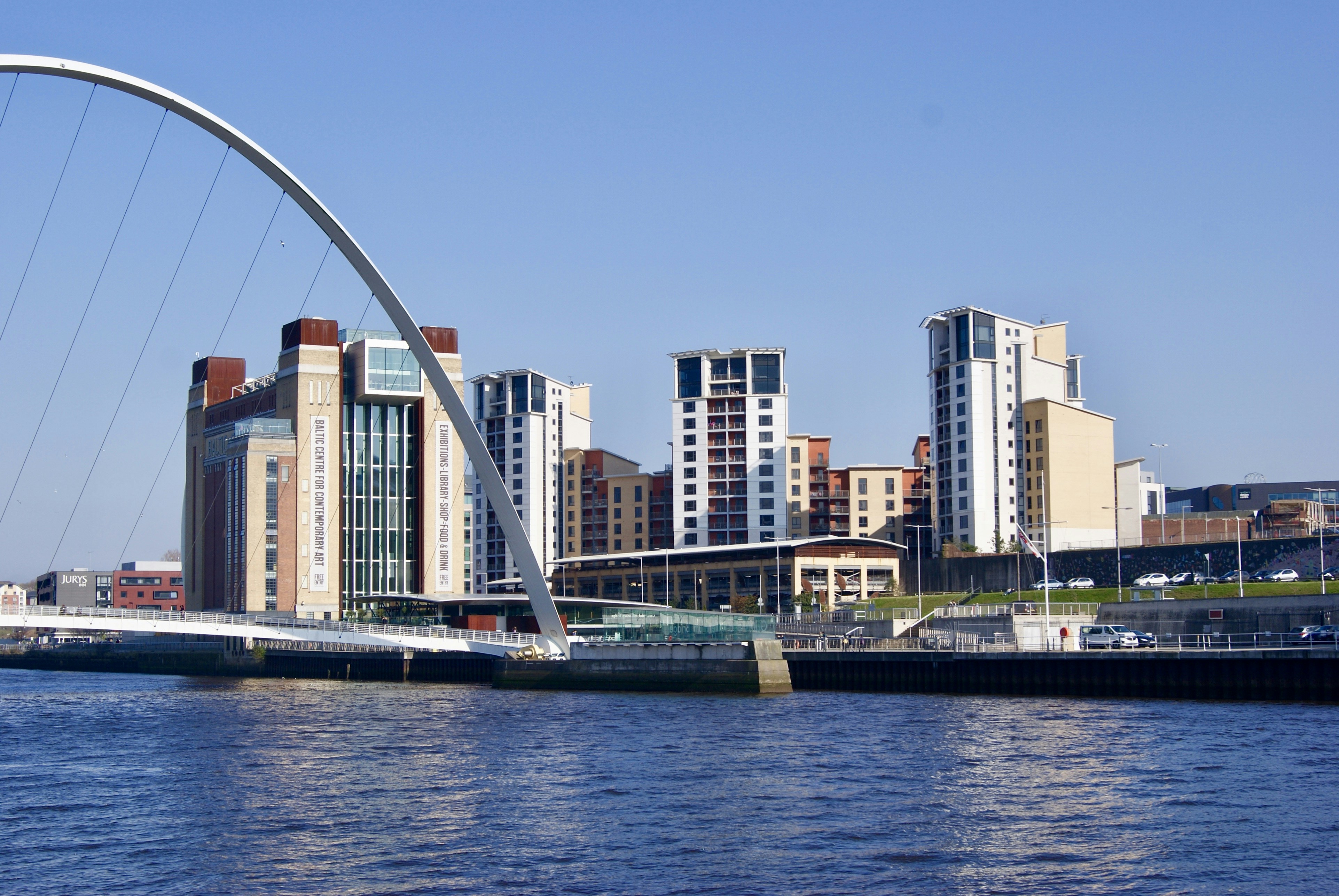 Contemporary buildings line the riverbank under a clear blue sky, with a distinctive arched bridge in the foreground.