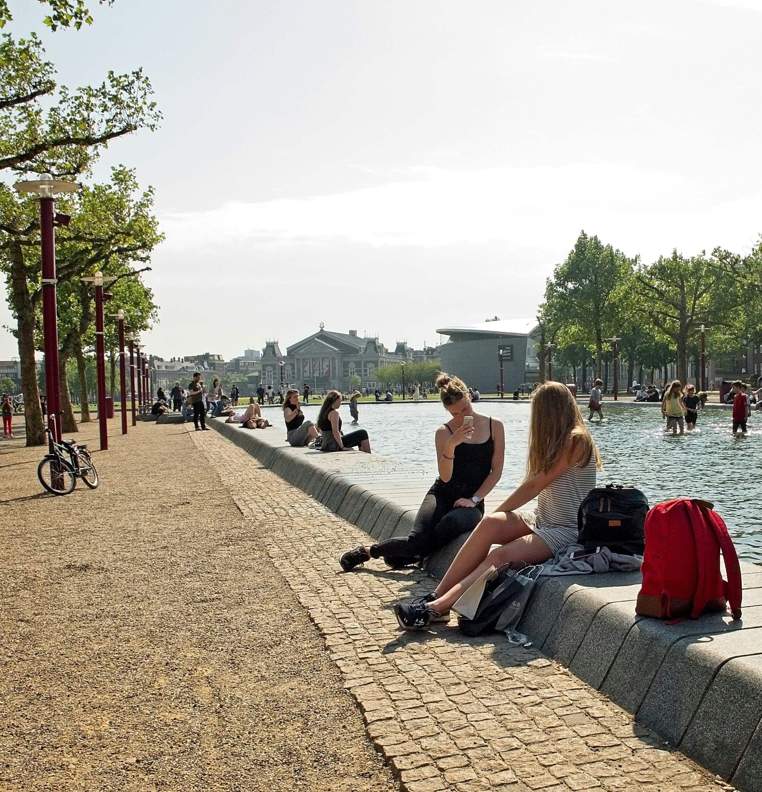 Two young women relax by a water feature in a vibrant urban park, surrounded by greenery and people enjoying the sunny day.