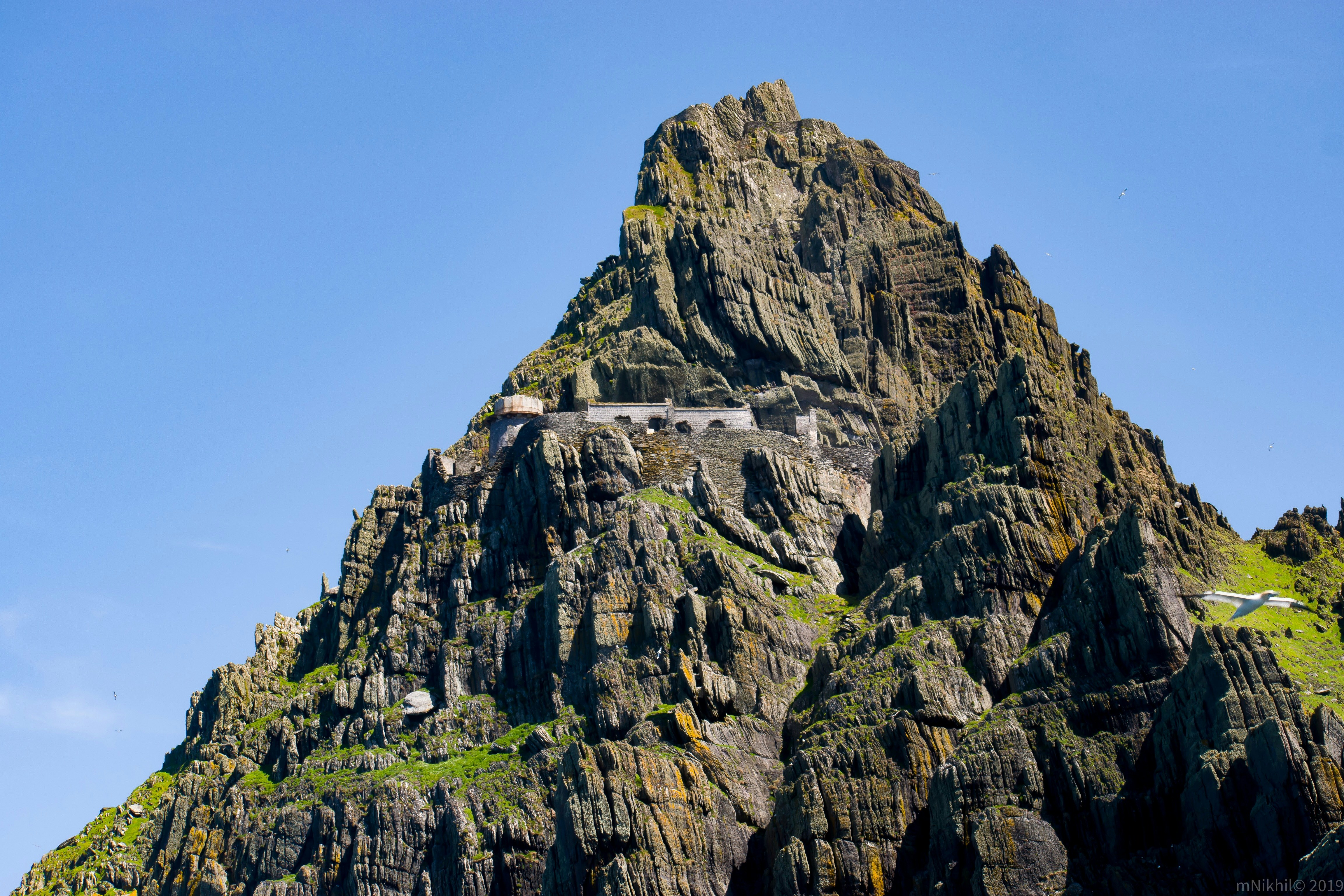 Ancient stone ruins perched atop a rugged mountain peak, surrounded by lush greenery against a clear blue sky.