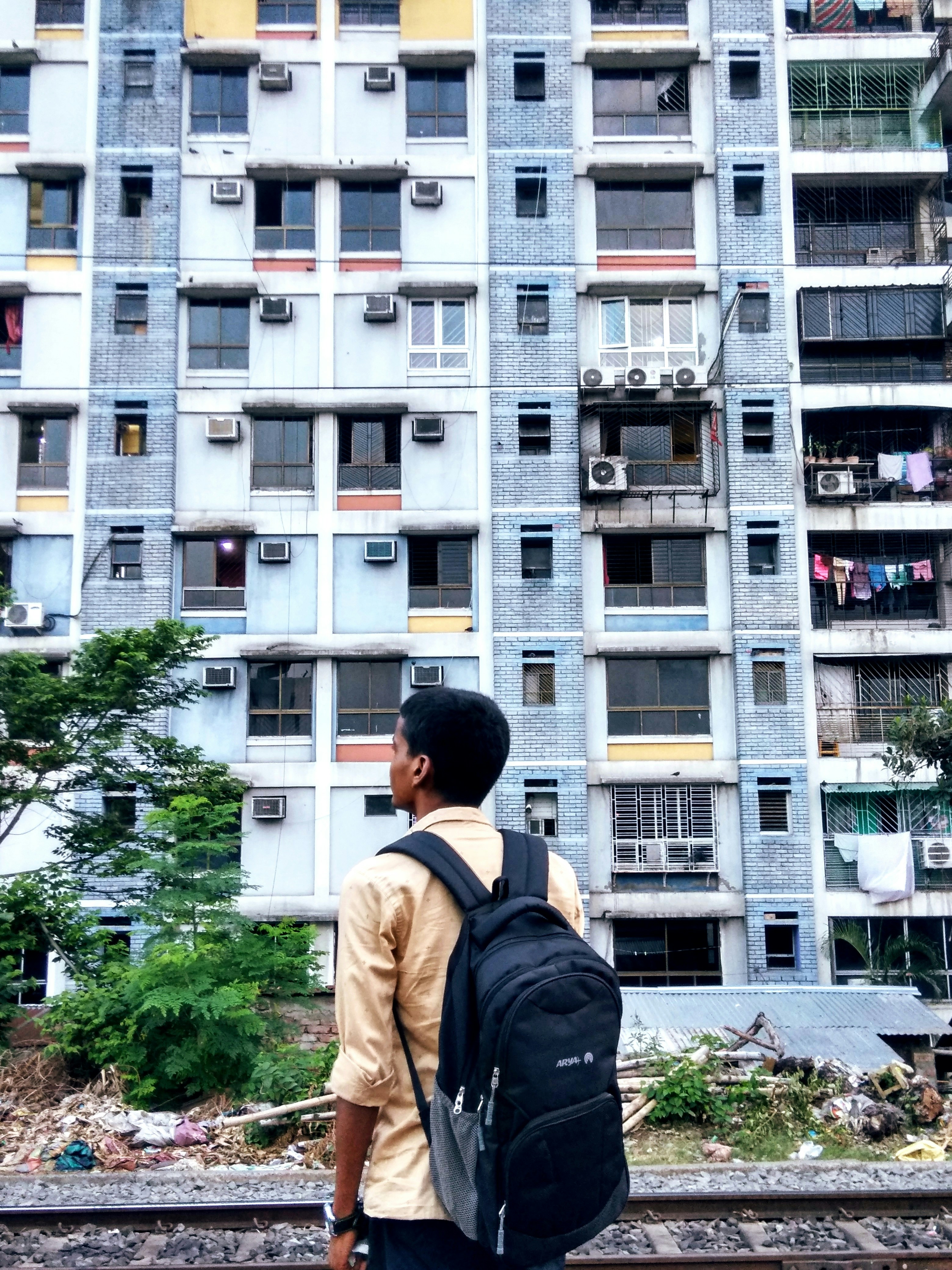 Street-level photograph of a man with a backpack standing before a tall apartment block; rows of windows, air conditioners and laundry create a geometric urban backdrop.