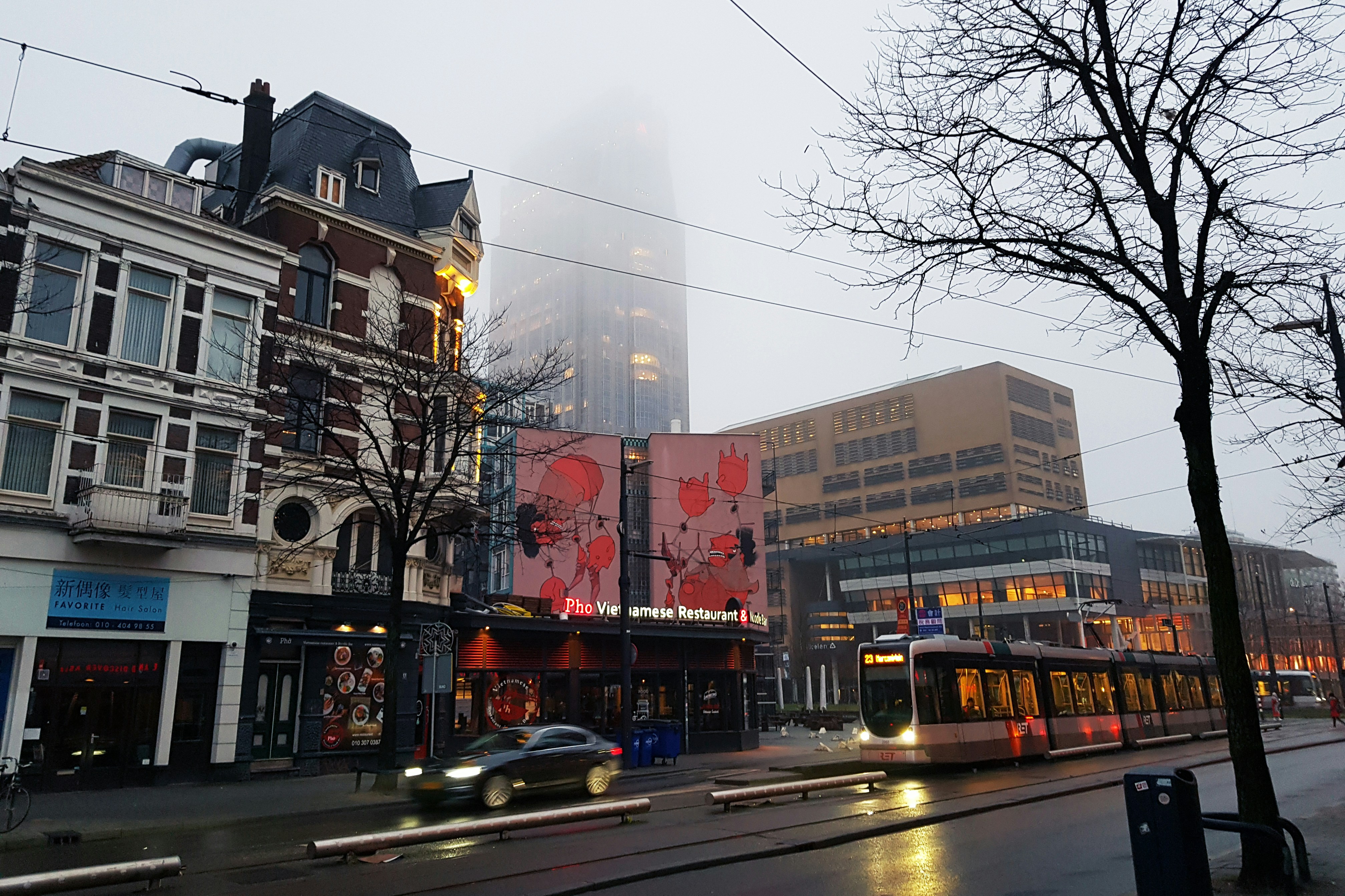 Foggy city street photograph featuring a tram and a bold red advertising billboard at the center, with a mix of historic and modern buildings. The moody scene emphasizes the rainy atmosphere and glowing city lights.