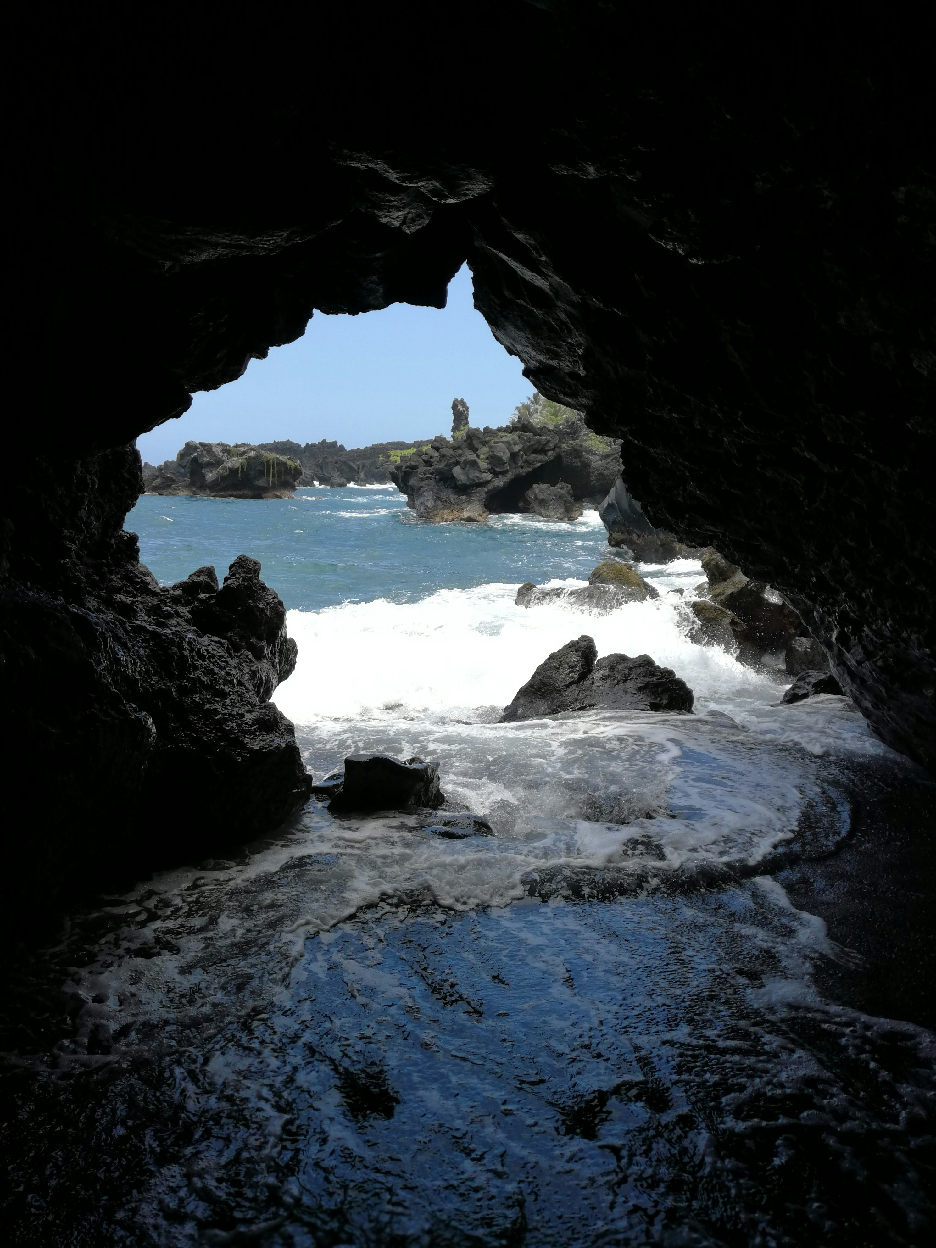 Dark coastal cave opens to a sunlit sea, jagged rocks and white foam framed by the arch against a blue sky.