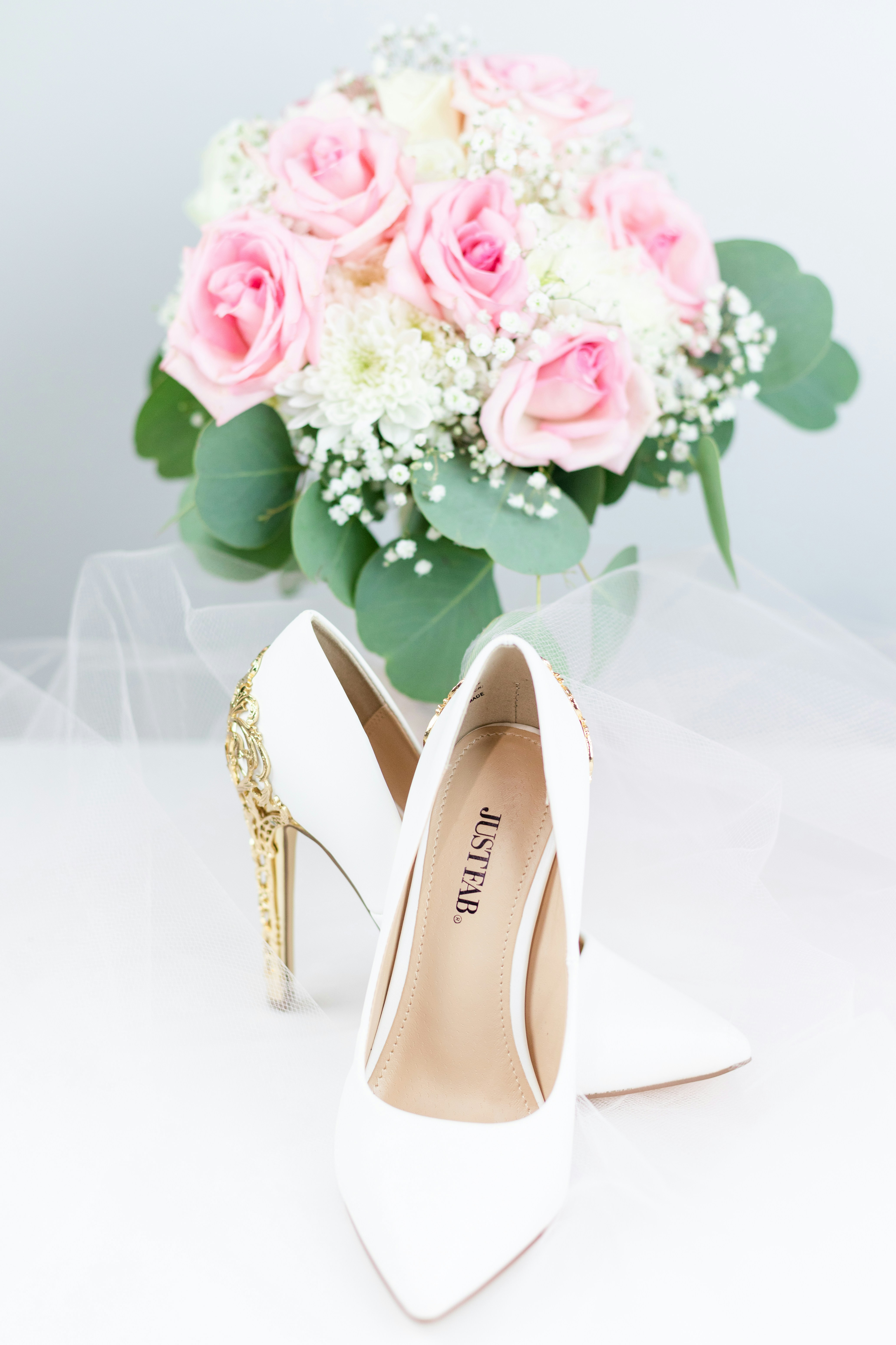White high-heeled shoes with intricate detailing positioned beside a bouquet of pink roses and delicate greenery.
