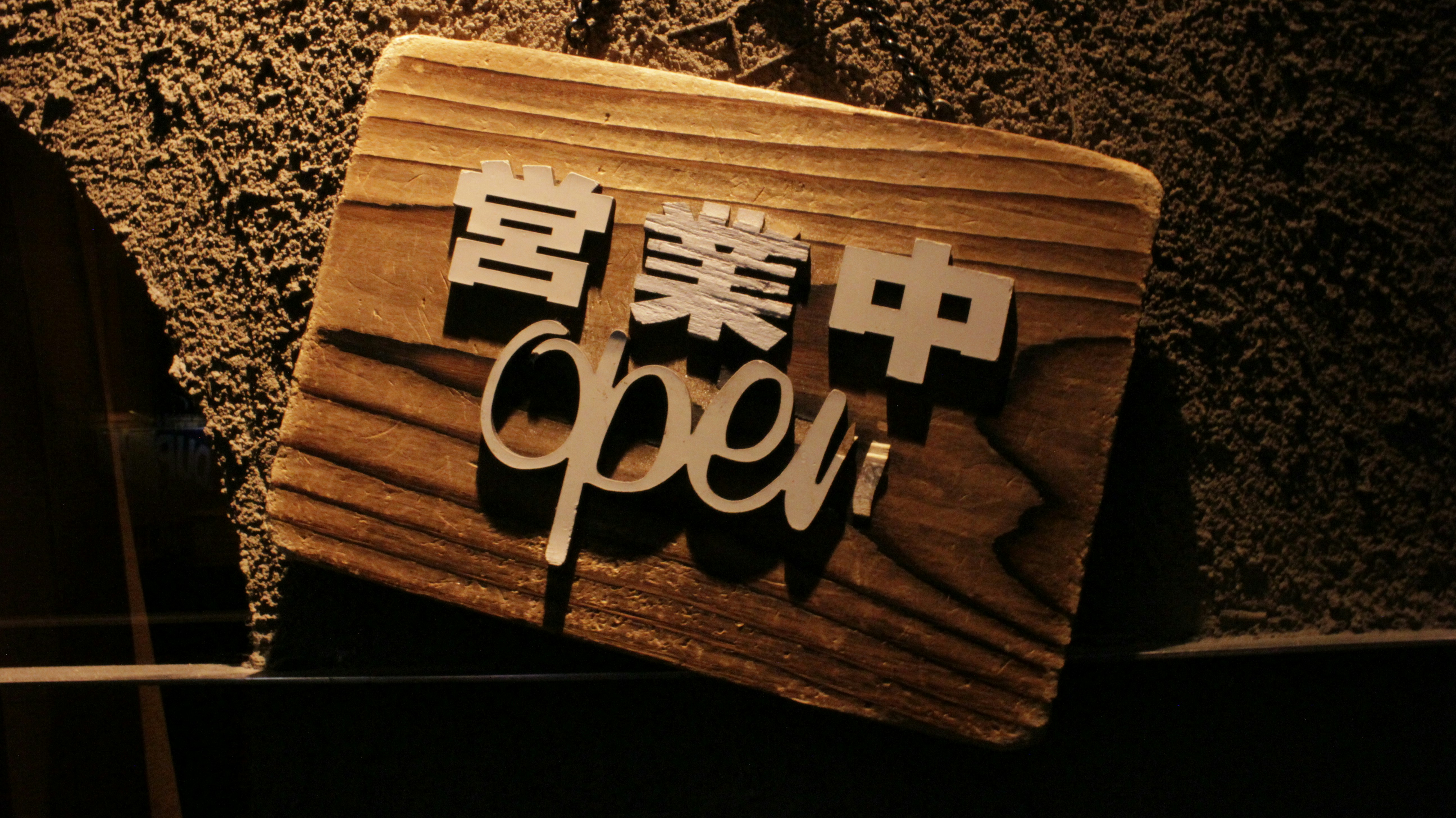Wooden sign with cut-out letters displaying 'open' and Japanese characters, illuminated against a textured wall.