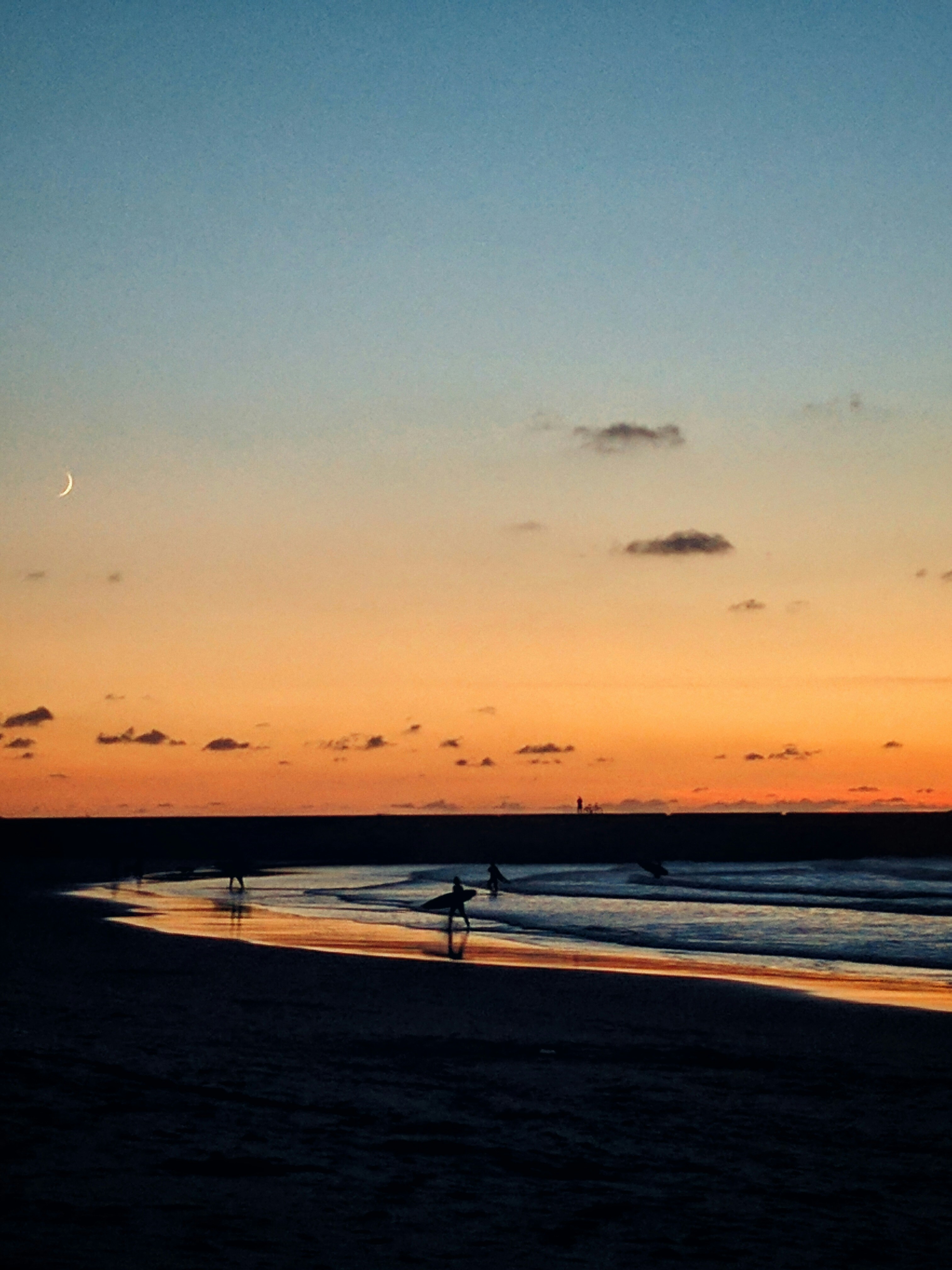 Surfers navigate gentle waves under a crescent moon at twilight, with vibrant hues of orange and blue in the sky.