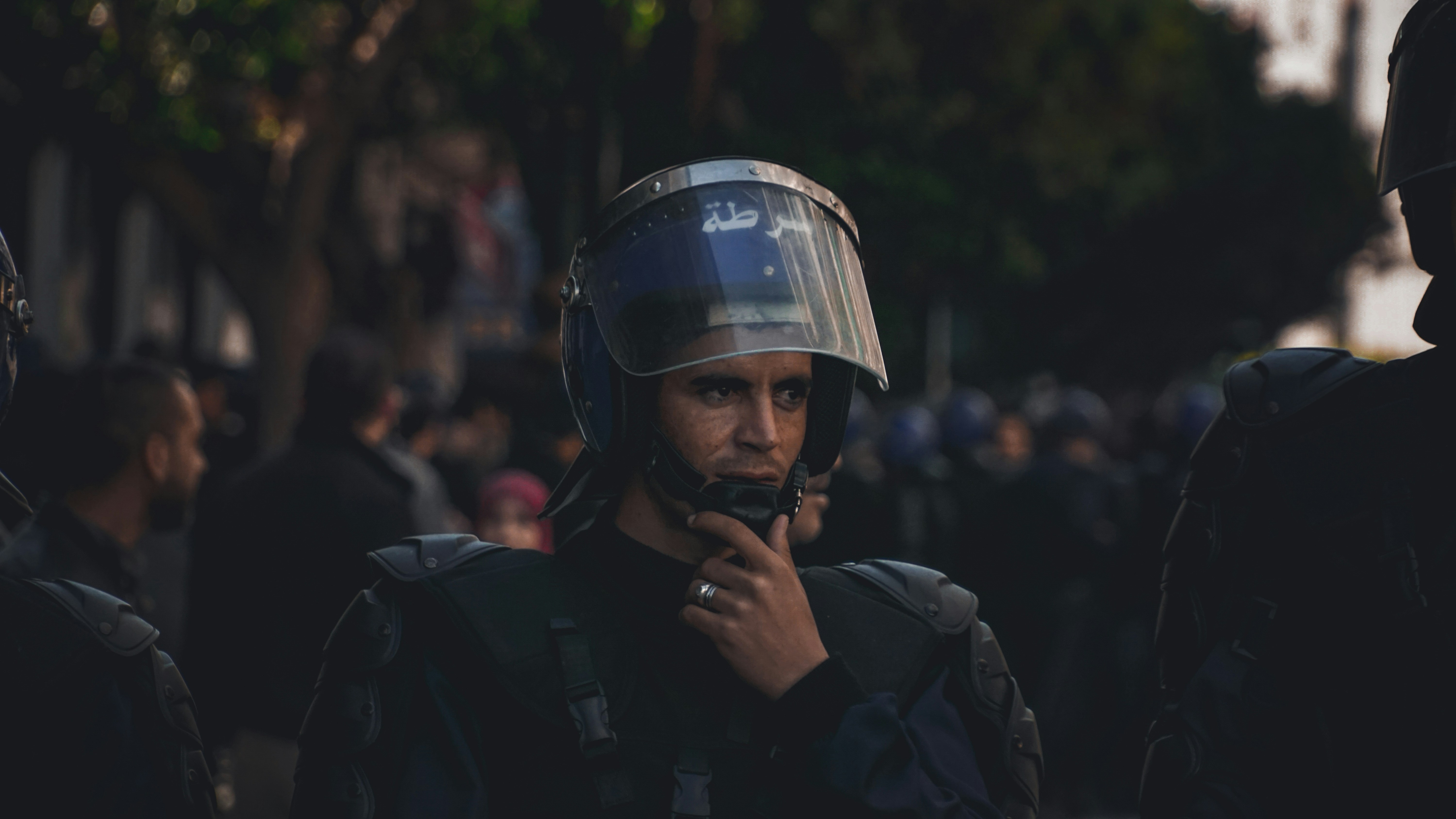 A contemplative police officer in riot gear stands amidst a crowd, deep in thought, as tensions rise in the background.