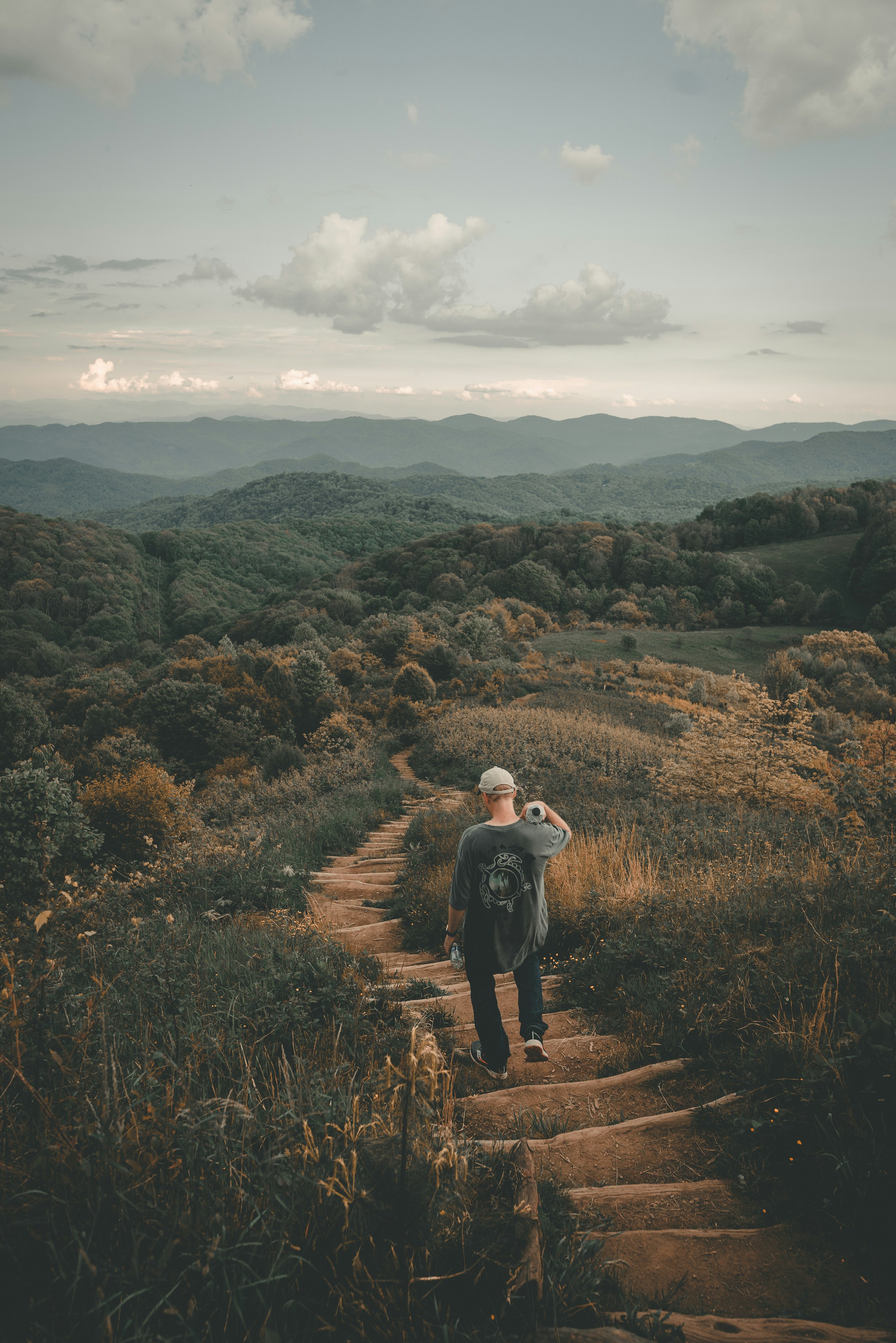 Hiker descending a rustic stone path surrounded by lush greenery and rolling hills under a cloudy sky.