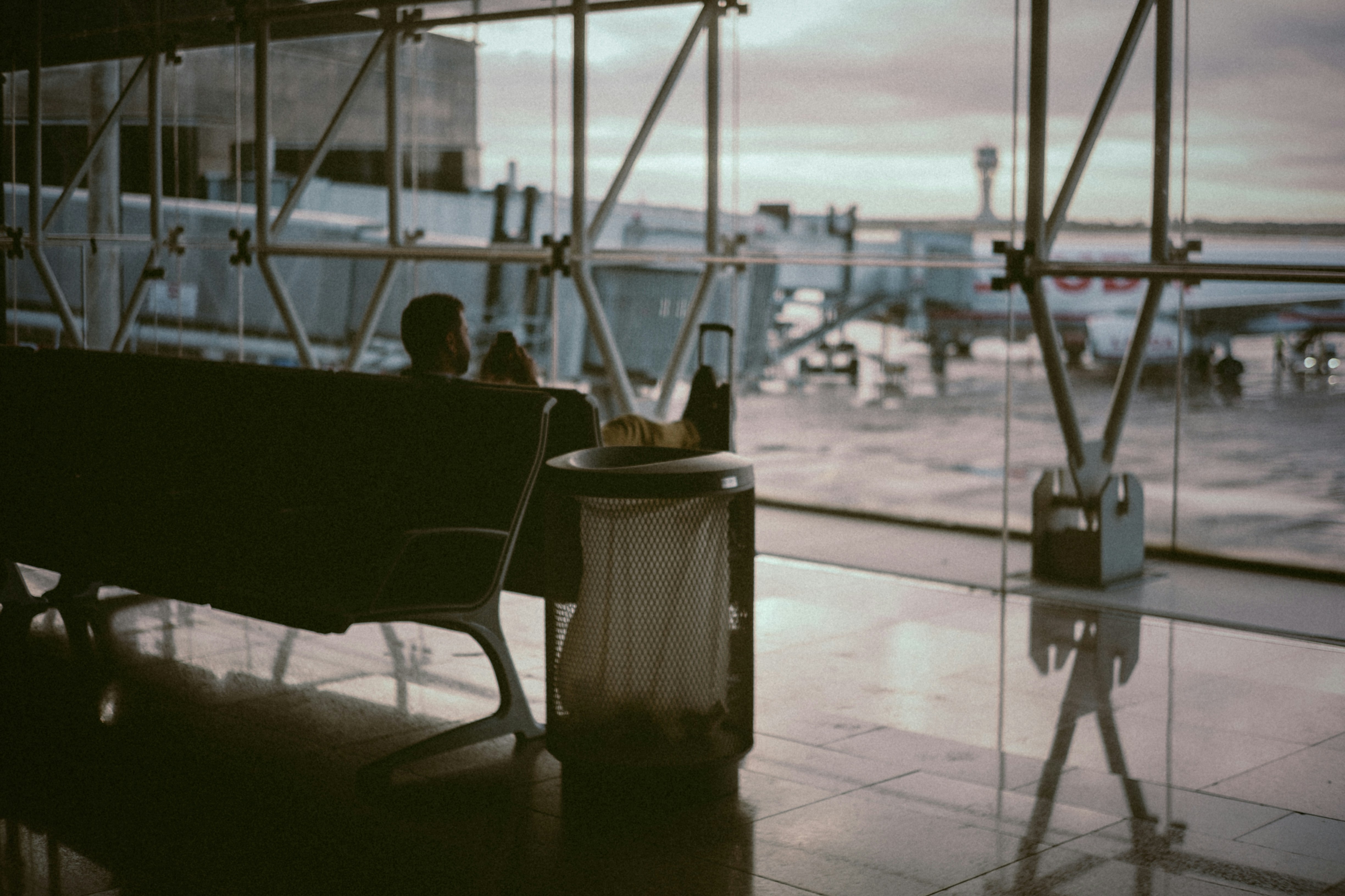 Passengers seated in an airport lounge, gazing out at the tarmac where planes await. A trash can subtly included in the foreground adds to the scene's realism.
