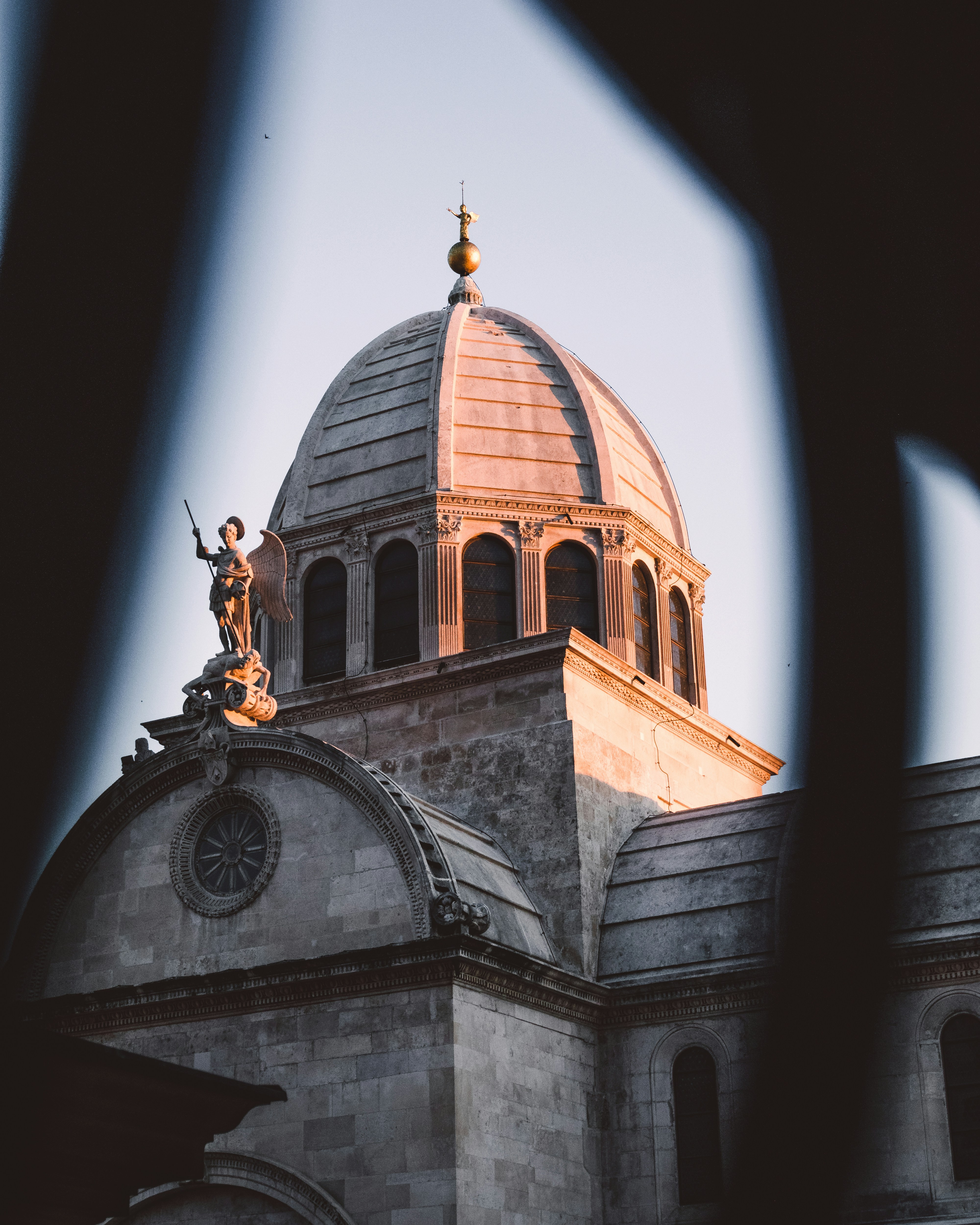 Statue of a guardian atop a dome, framed by architectural elements, during the golden hour.