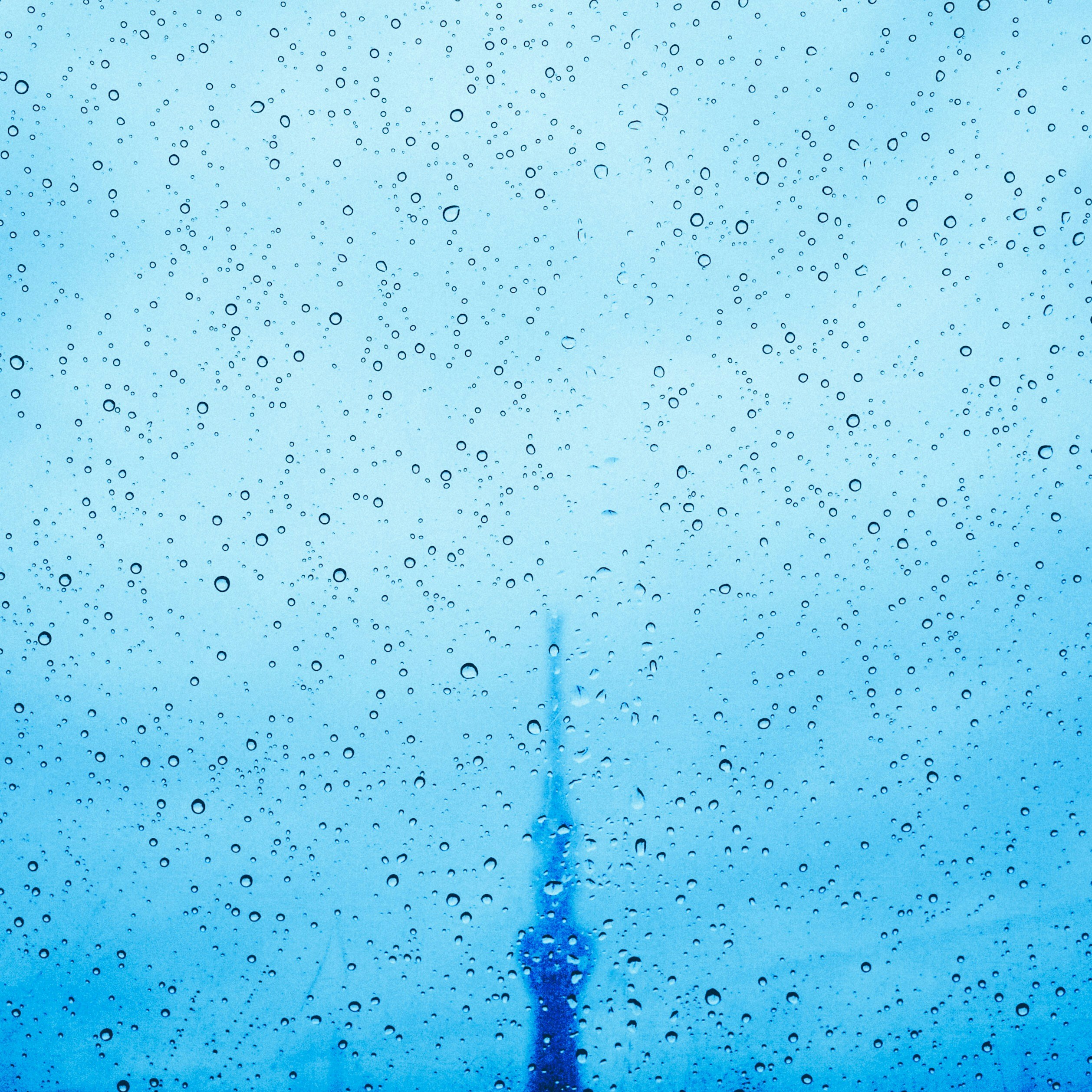 Raindrops on a glass surface obscure a distant city skyline, creating a serene, blurred effect. The scene evokes a sense of tranquility amidst urban life.