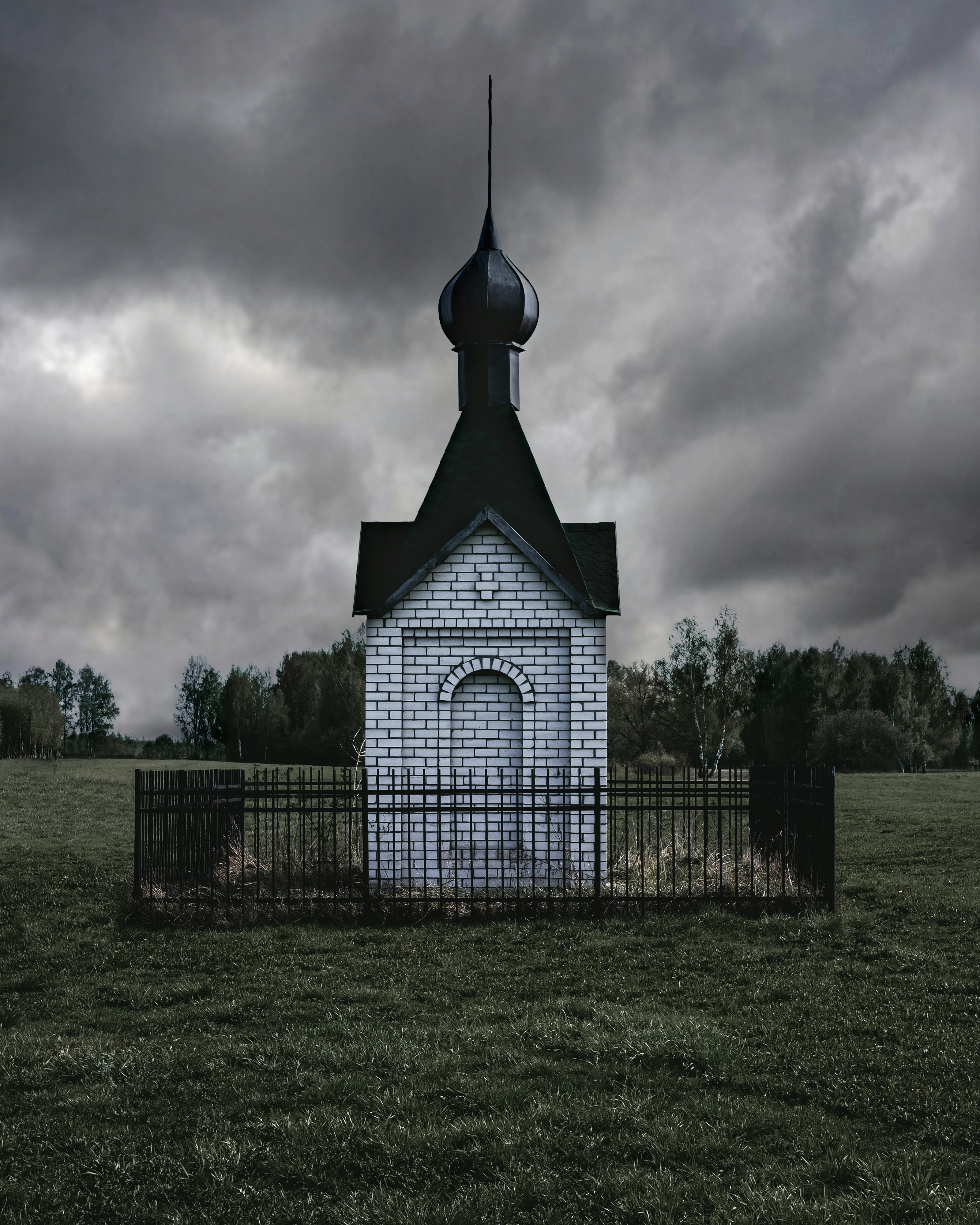 White brick chapel with onion dome and iron fence sits in an open field beneath a stormy, cloud-filled sky.
