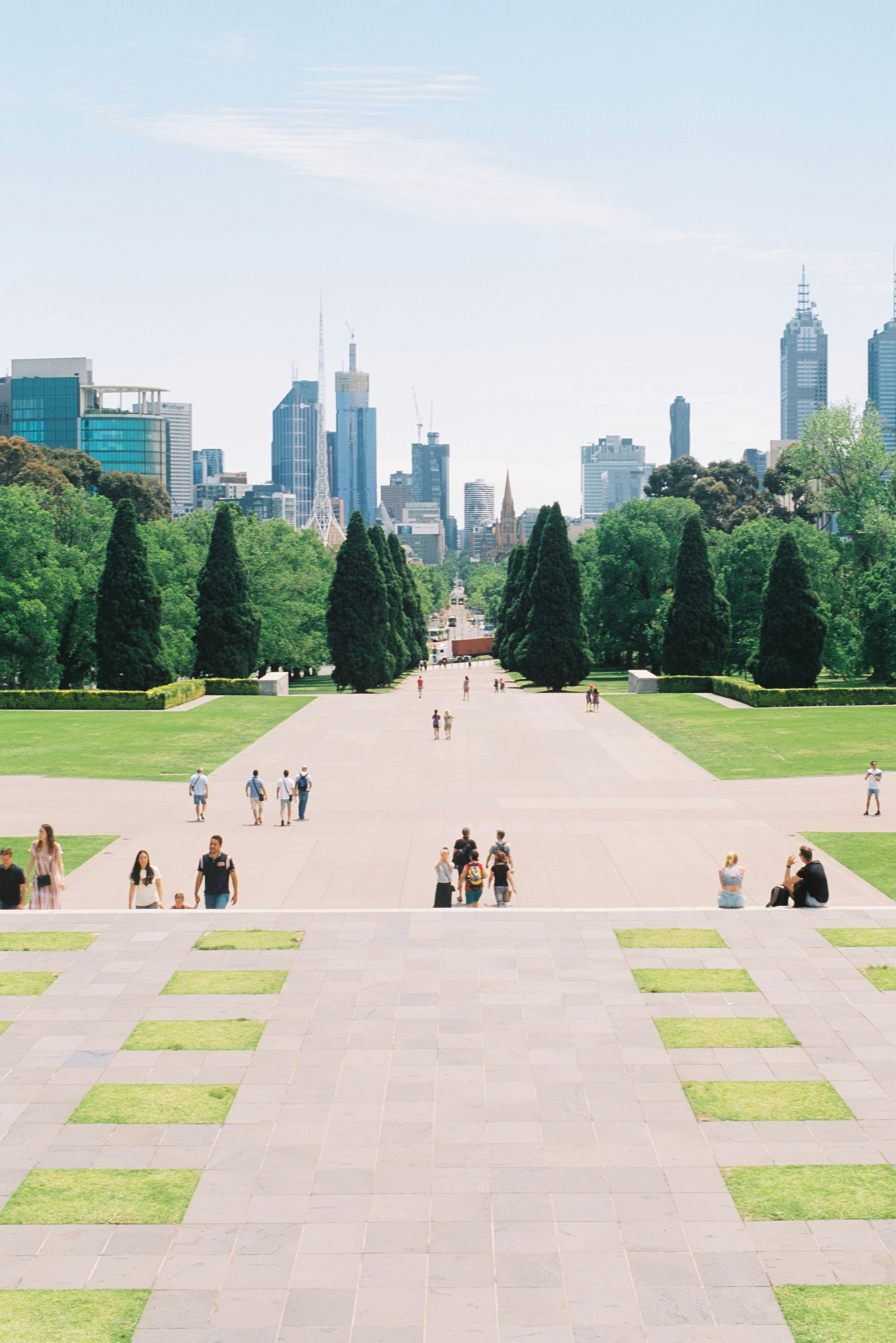Lush gardens lead the eye toward a city skyline, blending nature and urban life seamlessly. Visitors stroll along the pathways, creating a vibrant atmosphere.
