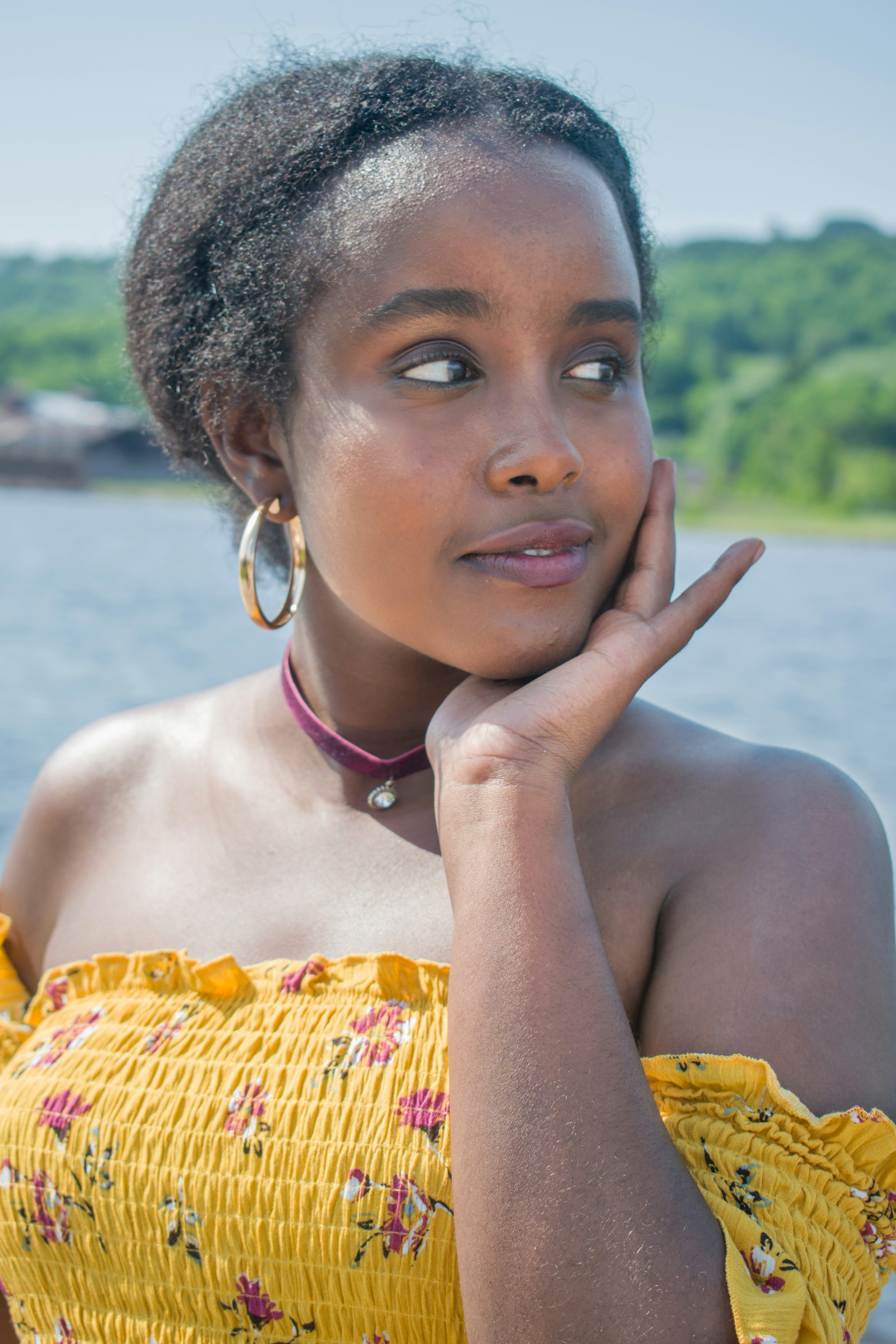 Young woman in a yellow floral top gazes thoughtfully with her hand on her cheek, set against a serene river backdrop.