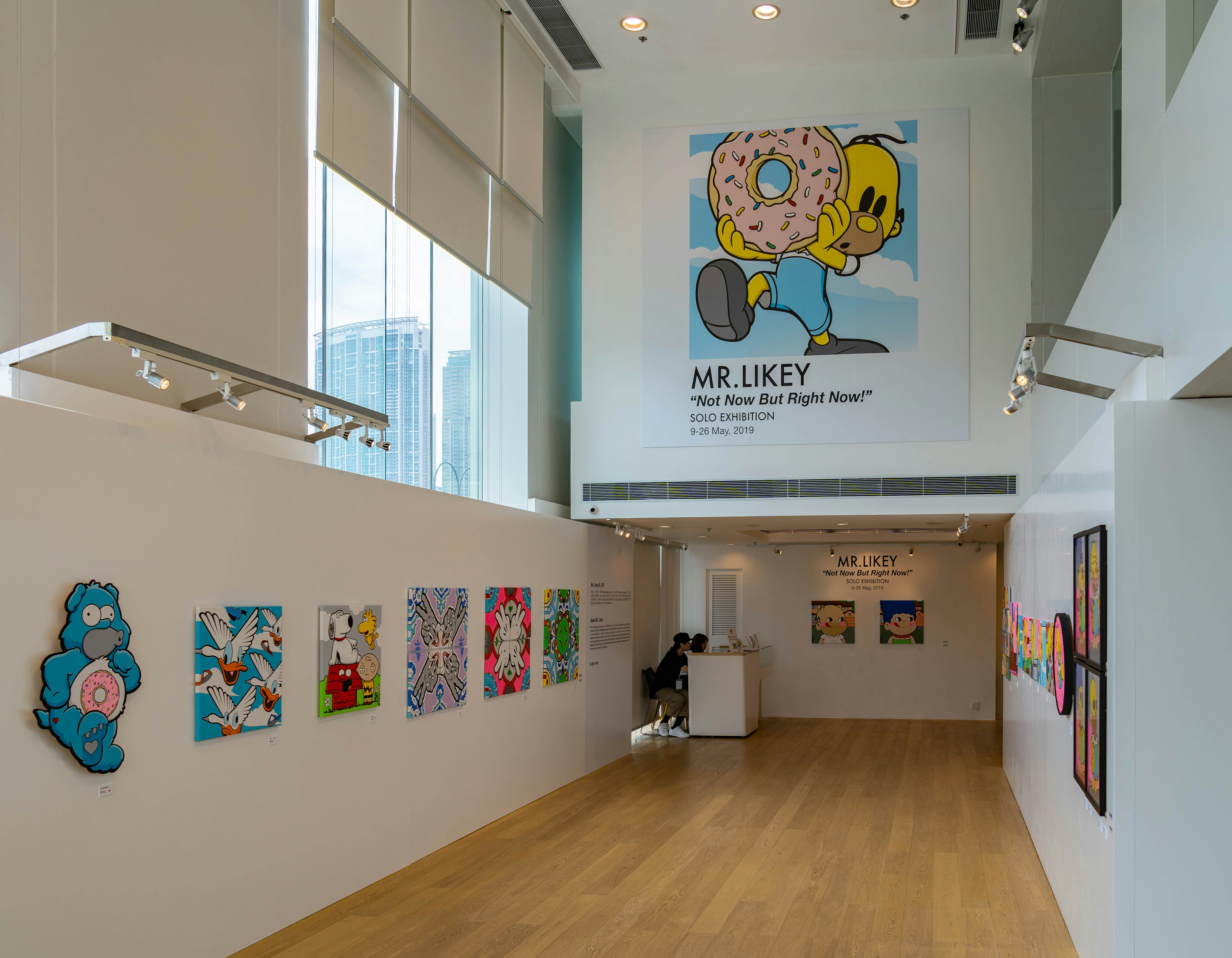 Art gallery featuring colorful pop art paintings and a large cartoon mural, with cityscape visible through tall windows.