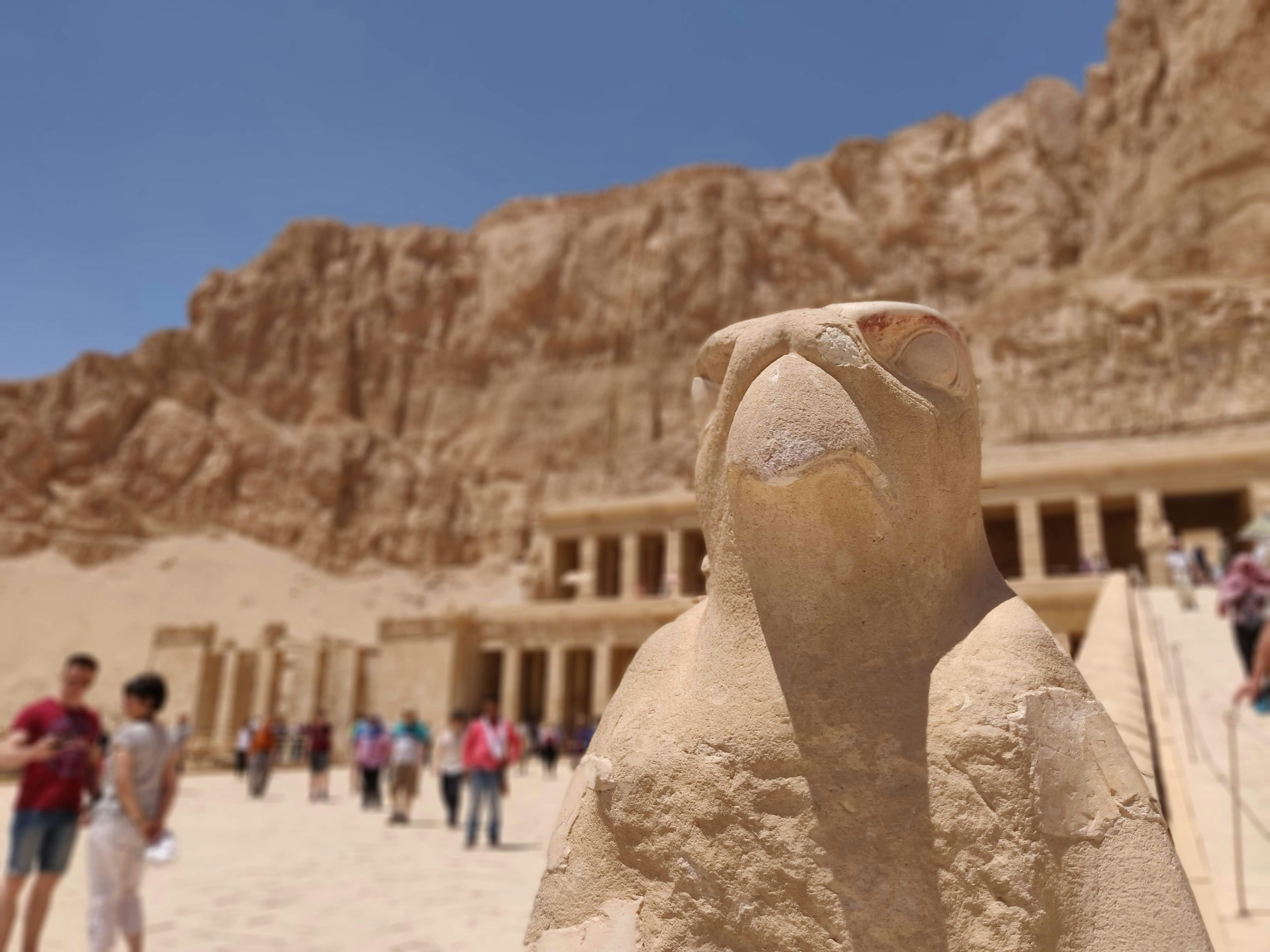 Stone statue of a falcon in front of ancient temple ruins, with visitors exploring the site in the background.