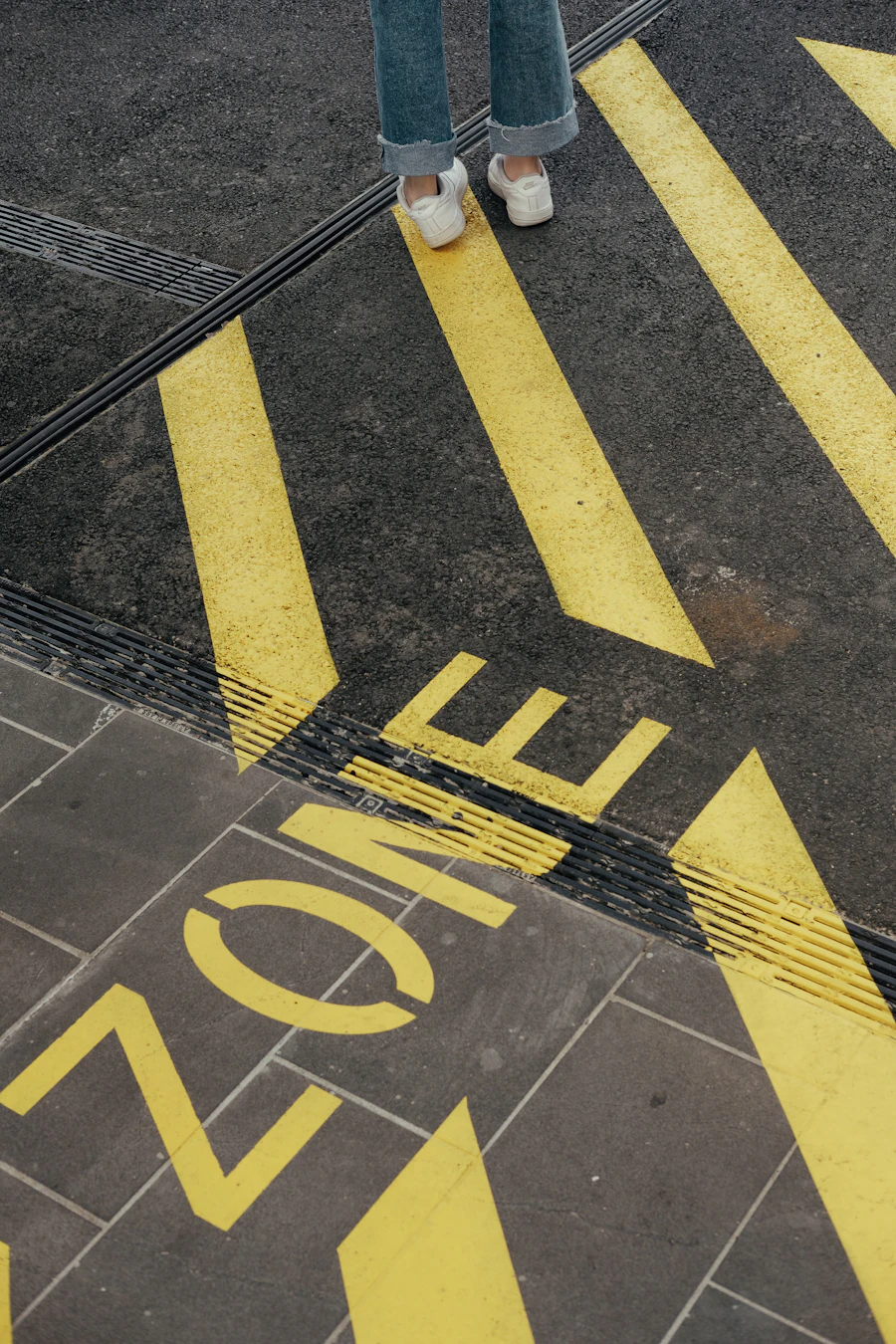 A person in cuffed jeans and white sneakers stands on dark pavement with large yellow directional markings and the word “KEEP” visible. Diagonal lines and drains create a graphic urban pattern.